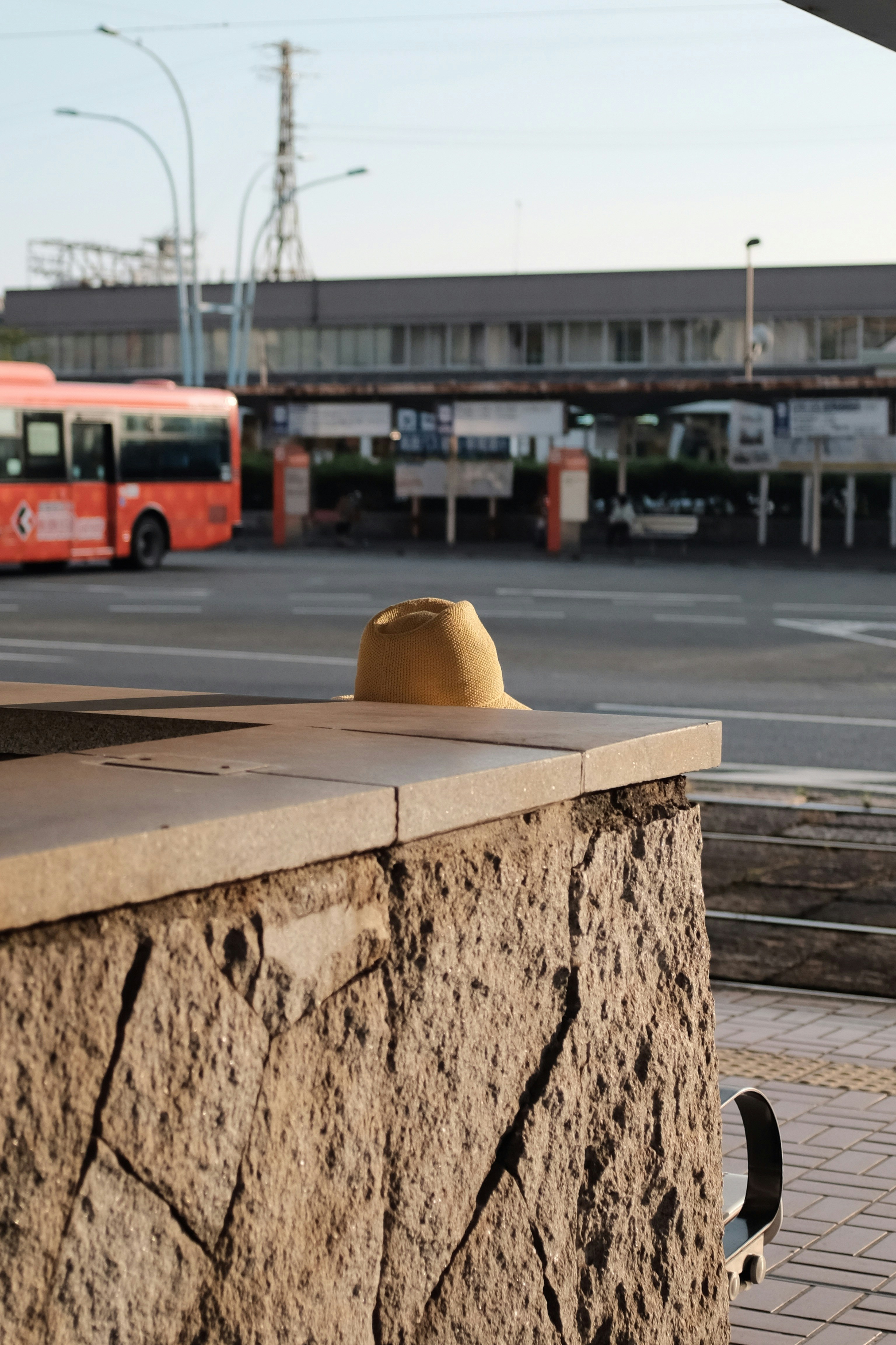 Yellow hat resting on a stone ledge at a bus stop, with a distant red bus and urban backdrop.