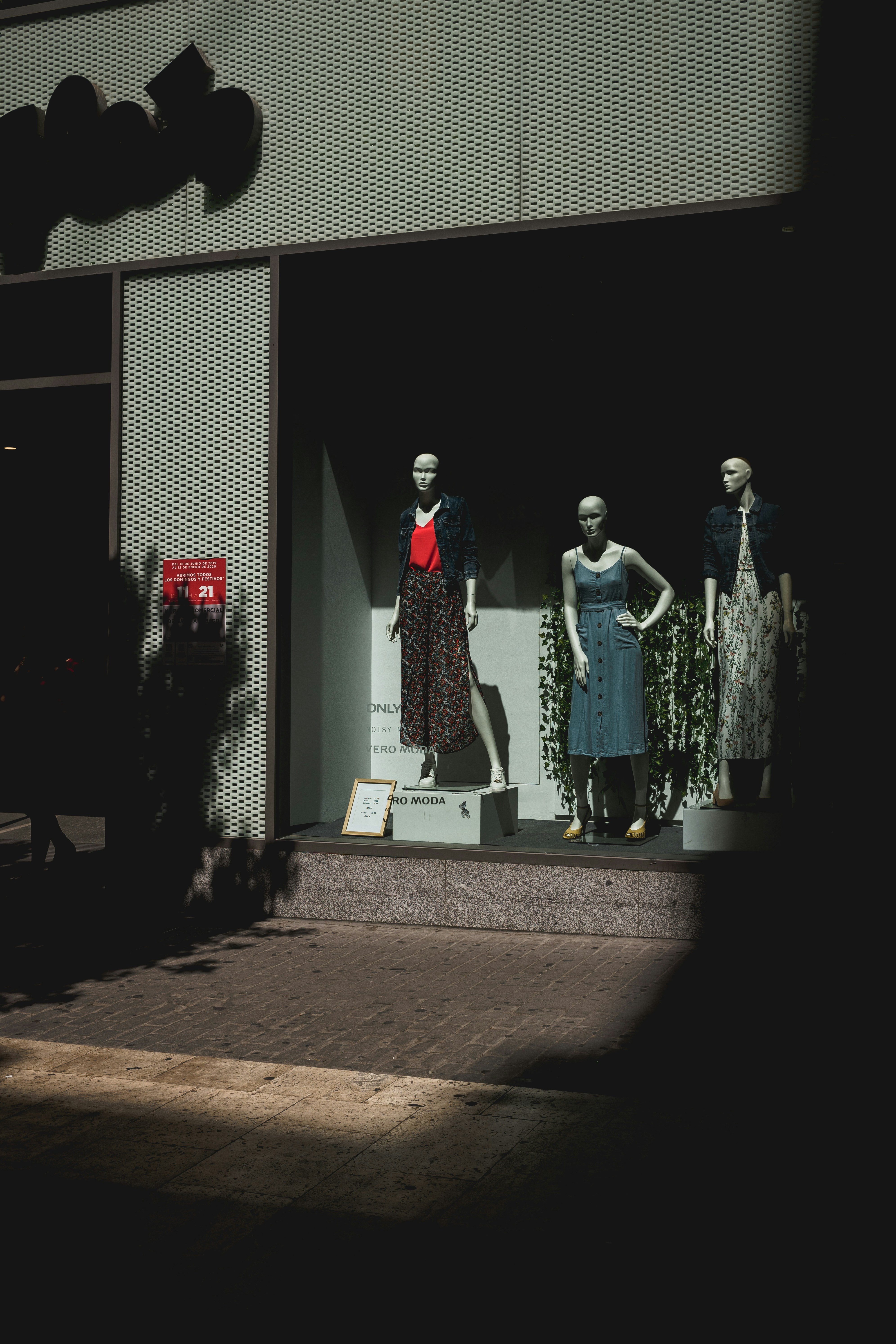 Three mannequins dressed in stylish outfits stand in a boutique window, framed by greenery and modern architecture.