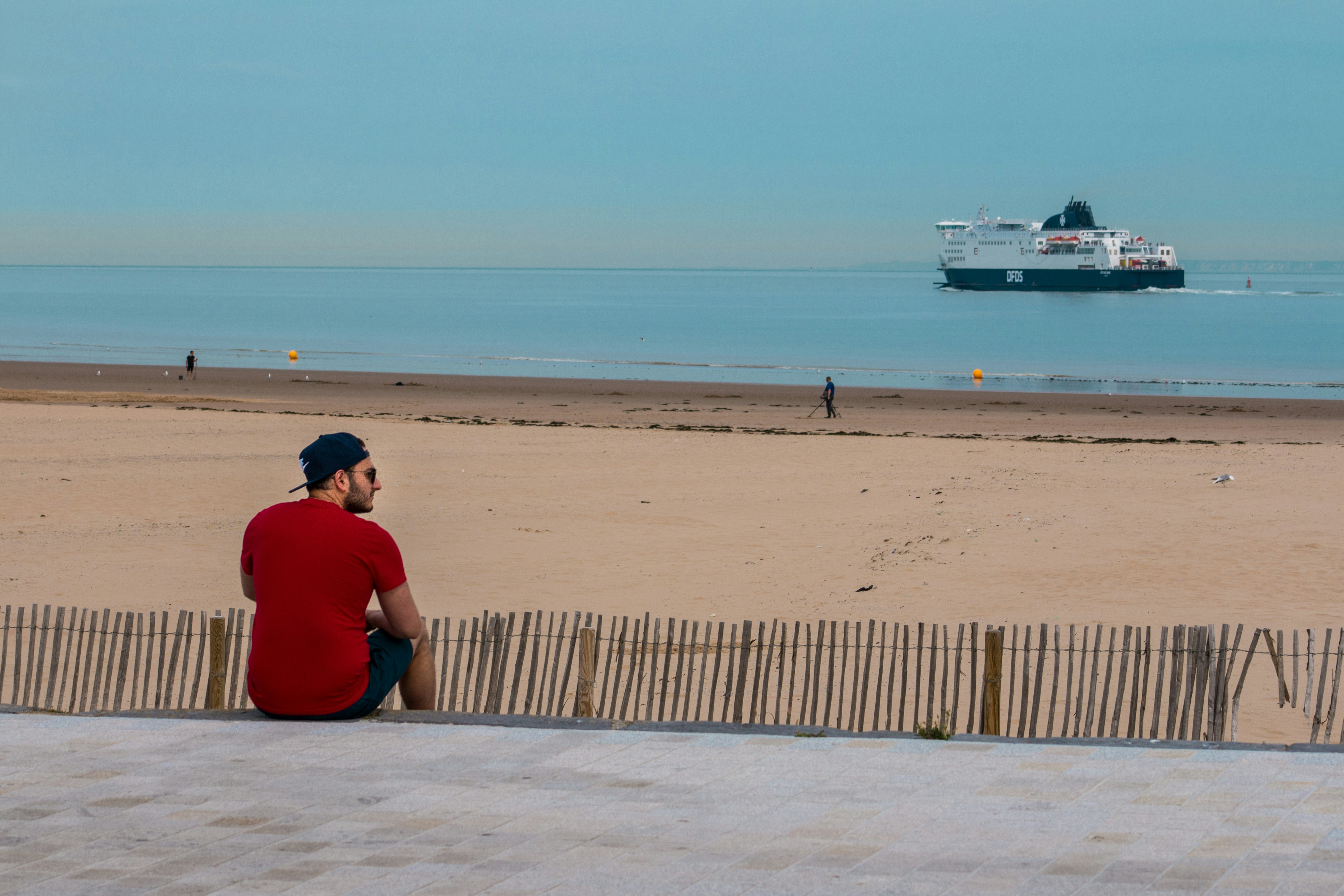 Man in a red shirt sitting on the beach, gazing at a ferry on the horizon.