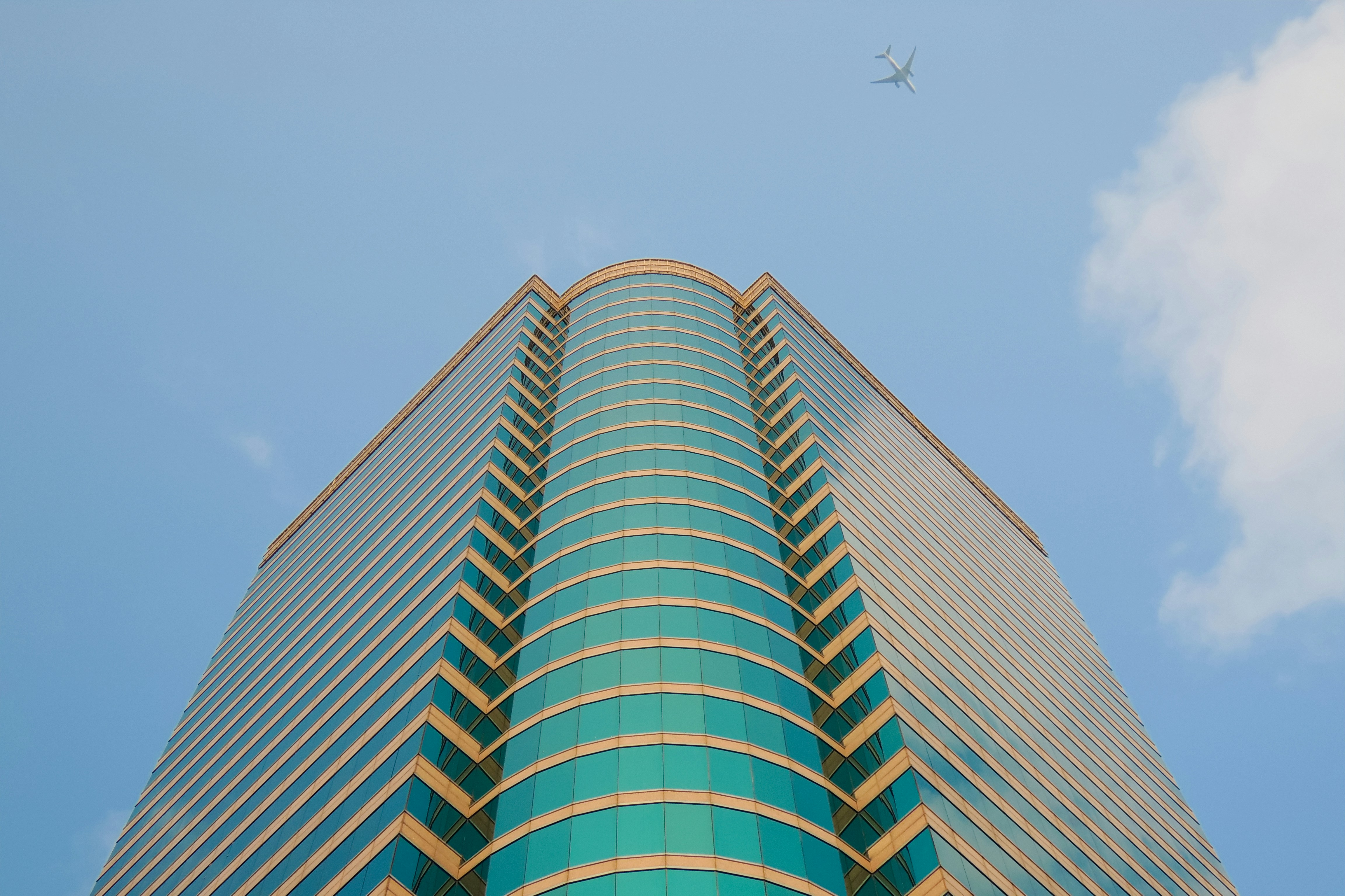 Modern glass skyscraper reaching towards the sky, with a plane flying above against a clear blue backdrop.