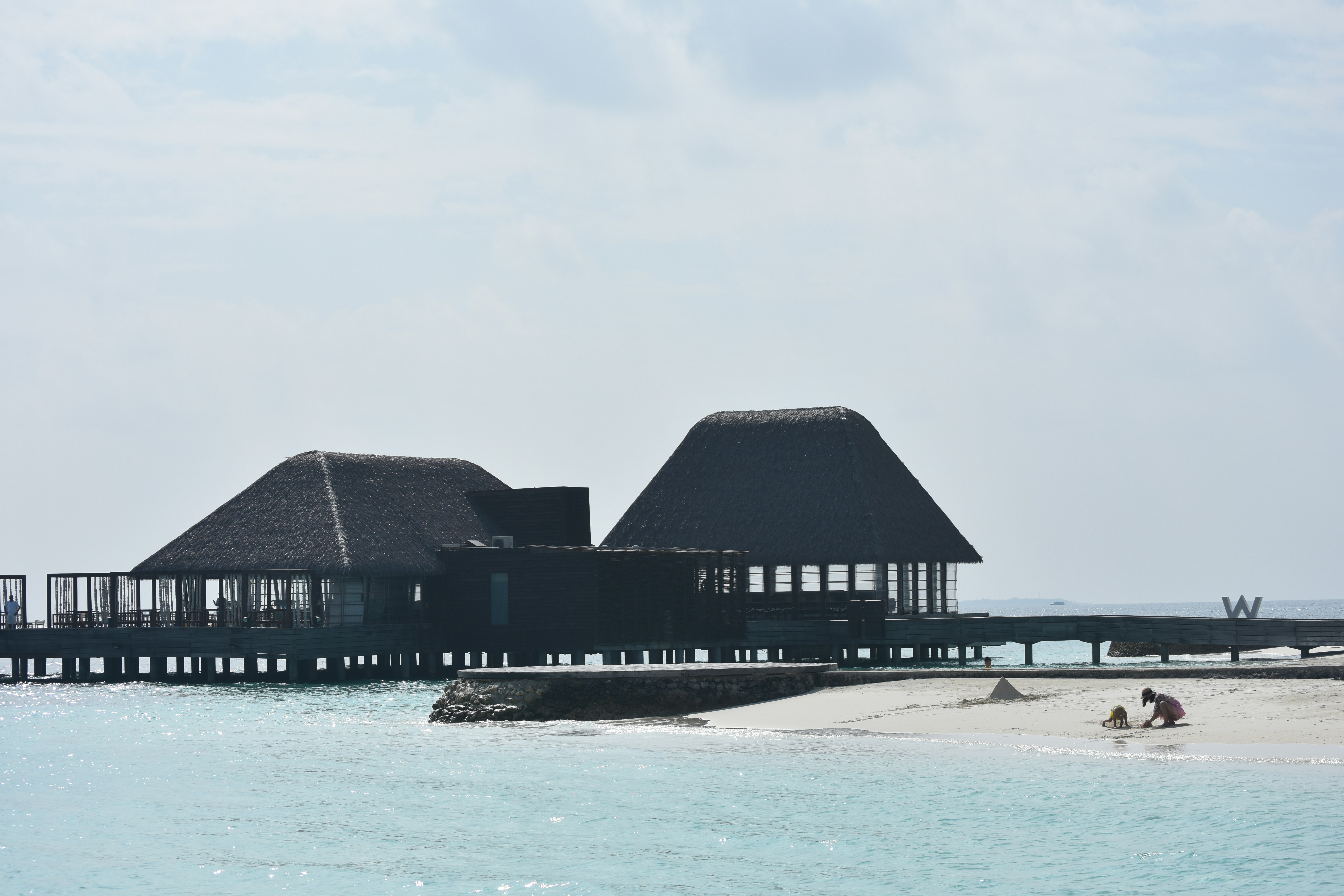 Tropical resort with thatched-roof villas overlooking calm turquoise waters, with beachgoers enjoying the sand nearby.