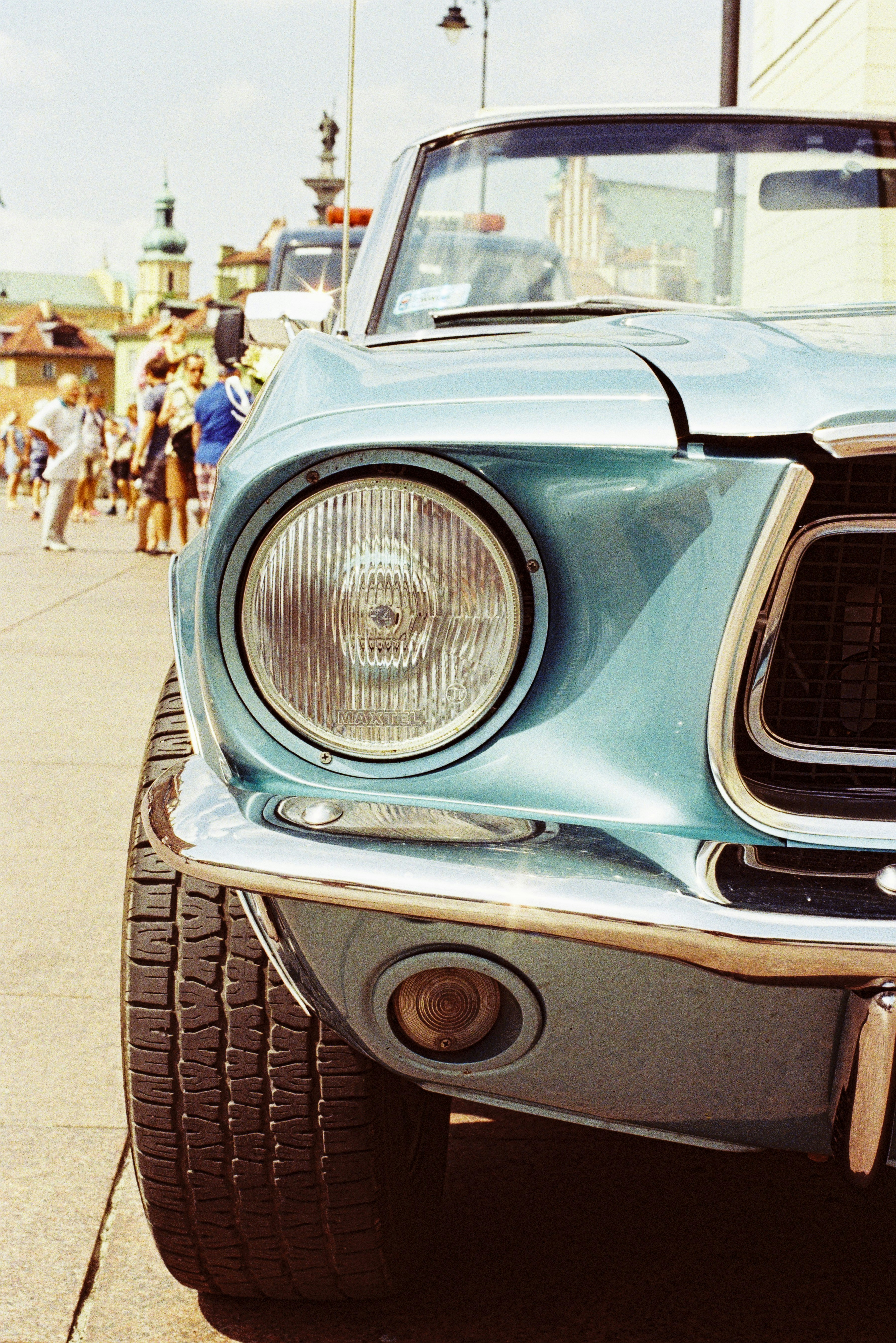Close-up of a vintage blue Mustang showcasing its iconic headlight and chrome detailing against a lively street backdrop.