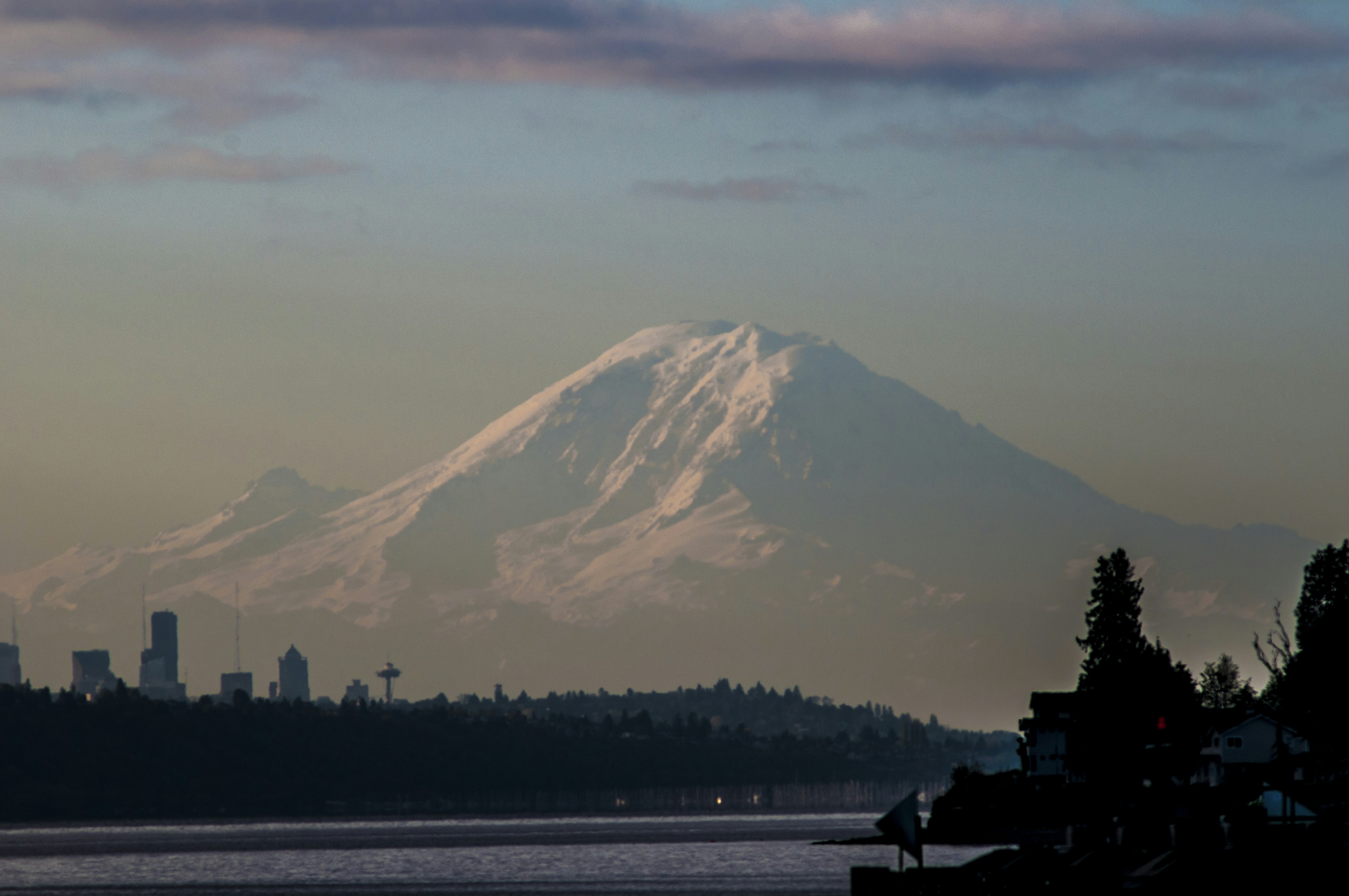 Snow-capped mountain under a pastel sky with a city skyline in the foreground at sunrise.