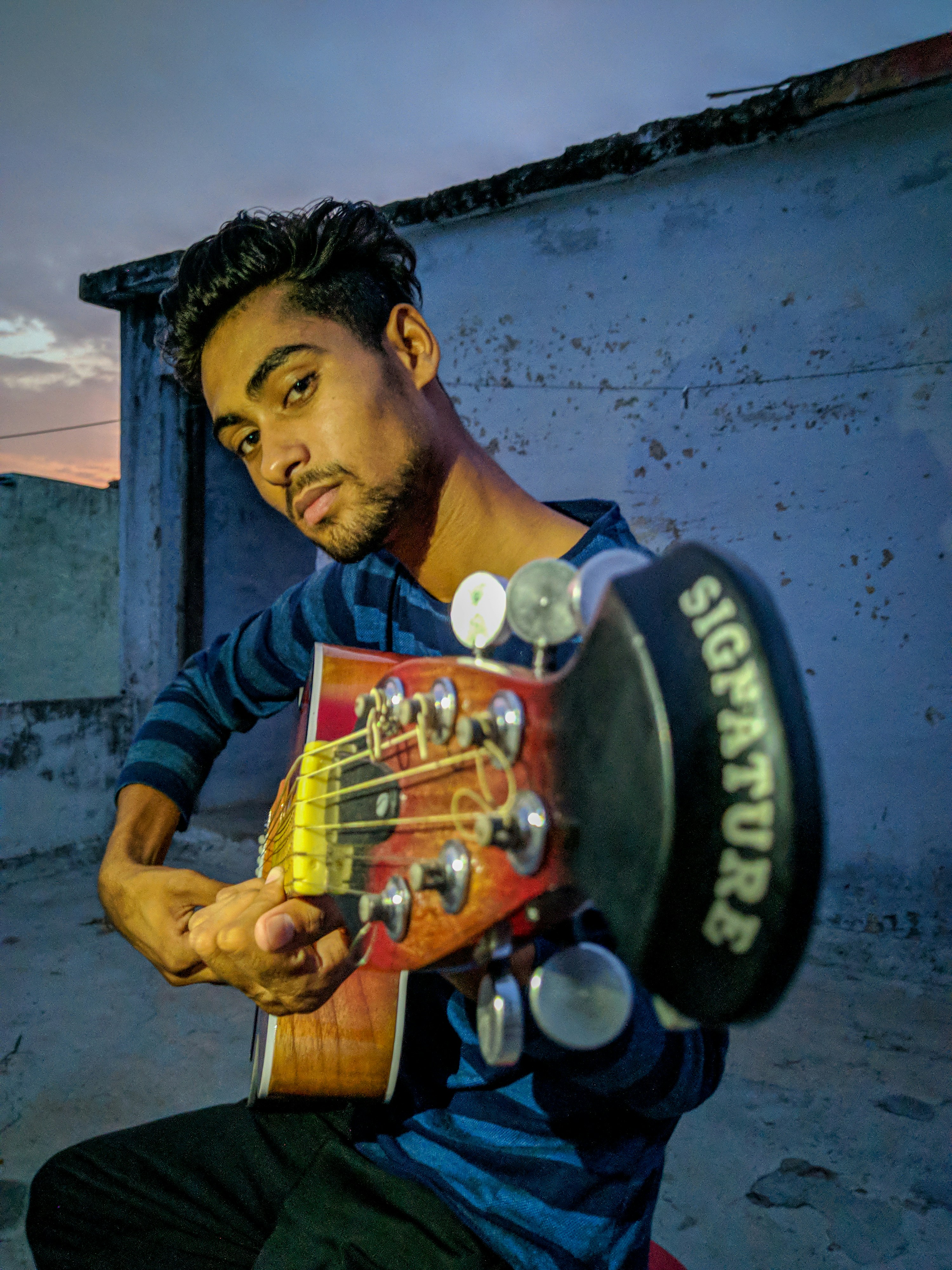 A man on a rooftop strums an acoustic guitar during blue hour, with cool ambient light shaping his features against a weathered wall.