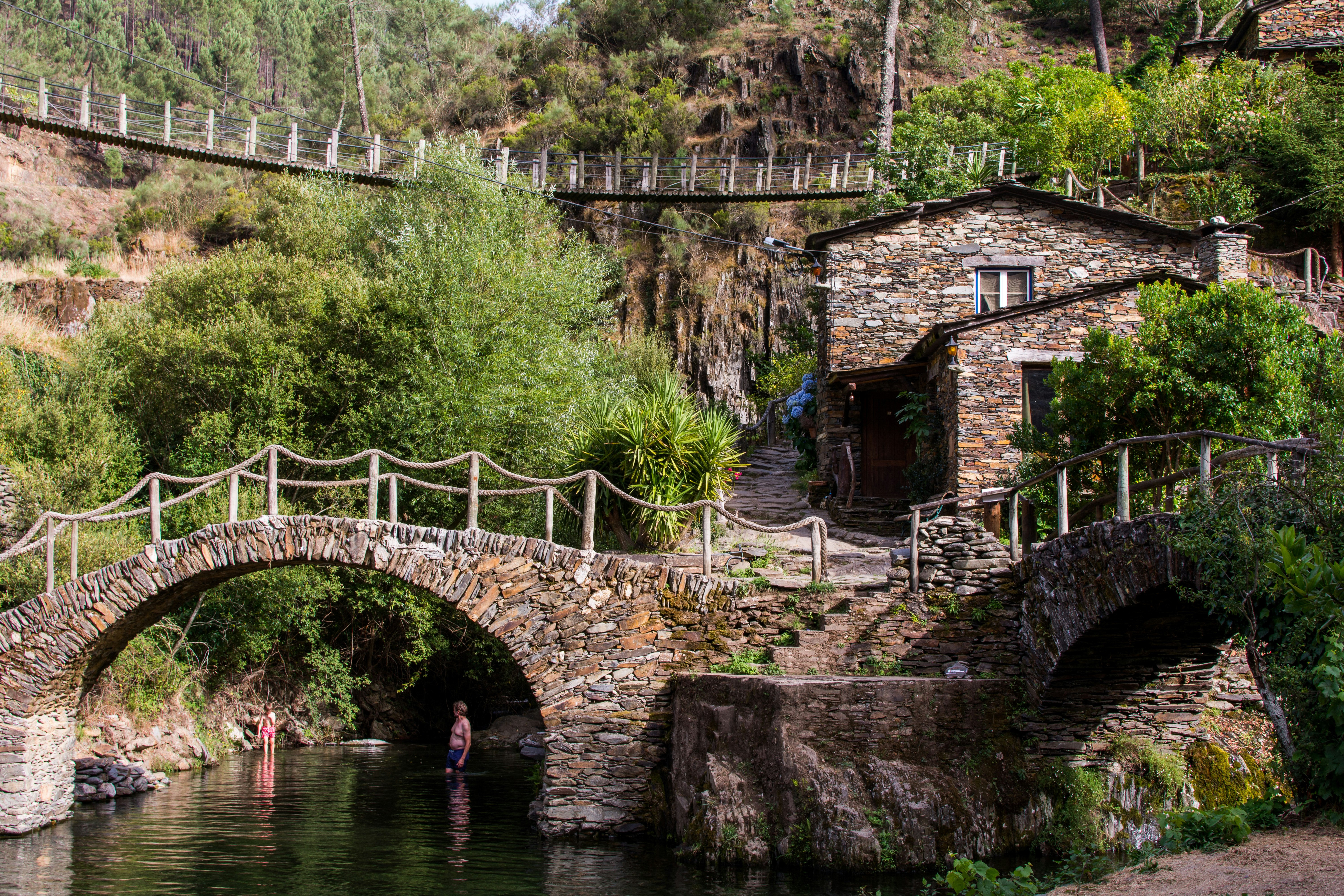 Stone bridge arches over a serene river beside a rustic stone house, surrounded by lush greenery.