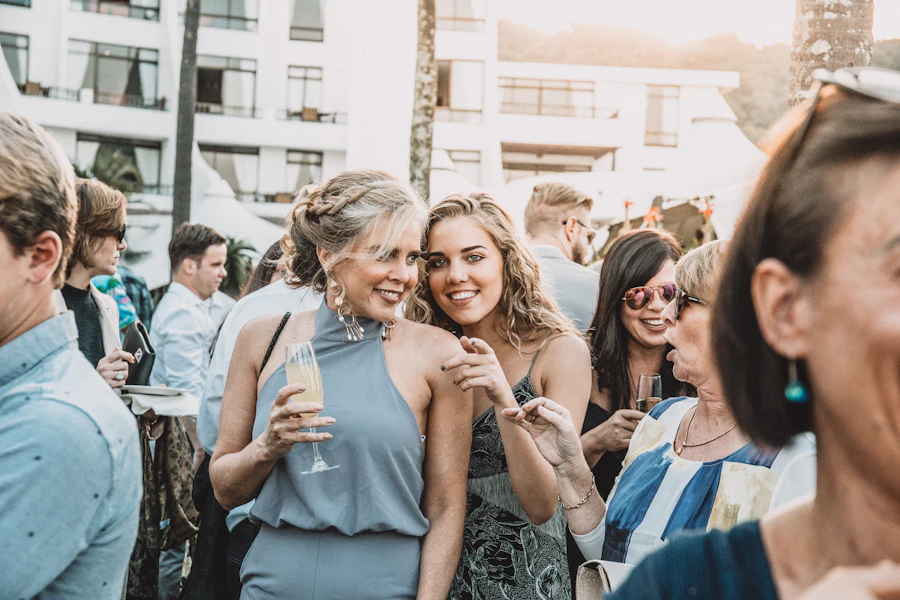 A group of well-dressed people socialize at an outdoor party, with two women smiling near the center and holding drinks. Bright buildings and palm trees frame the lively scene.