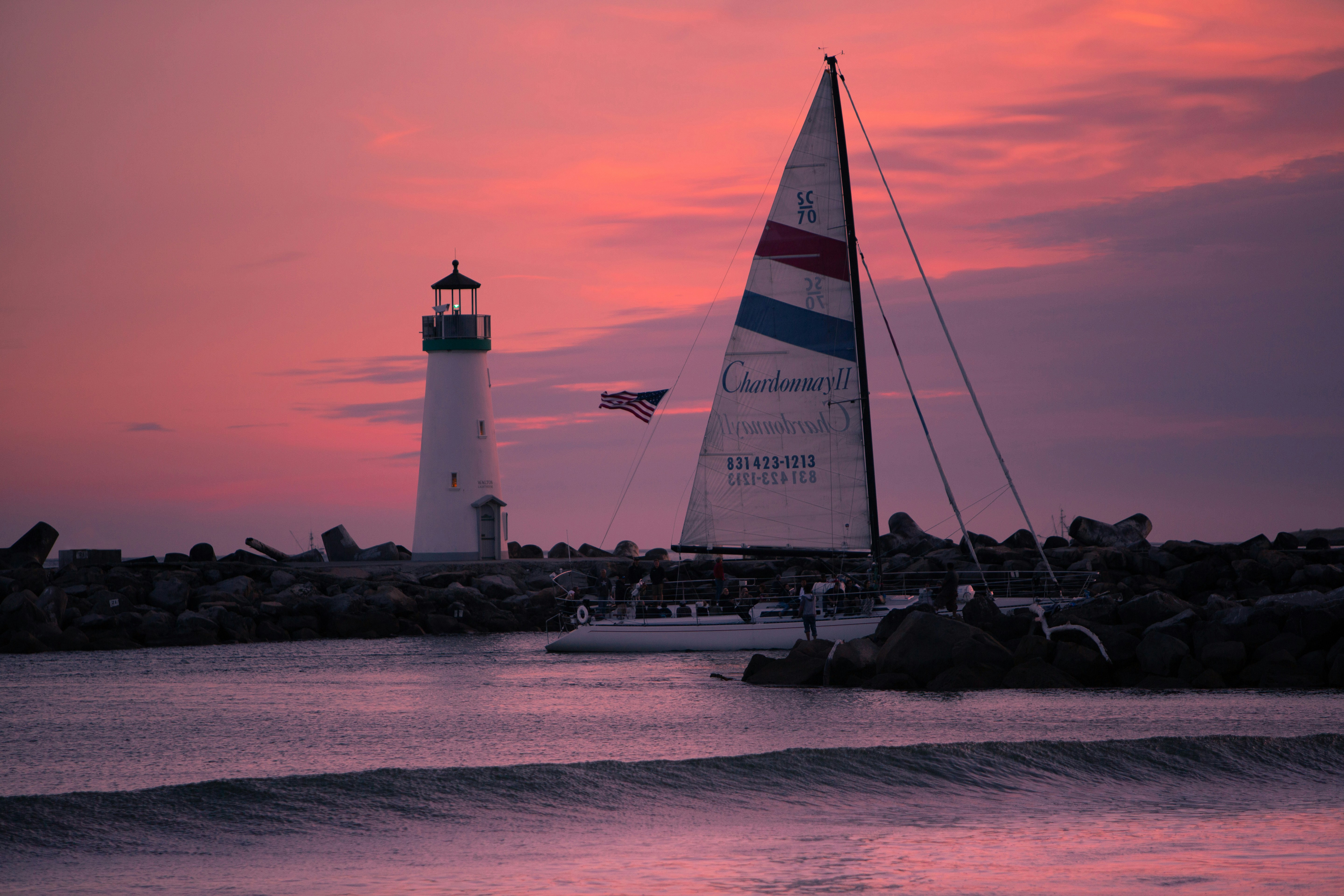 Sailboat gliding past a lighthouse under a vibrant pink and purple sunset sky.