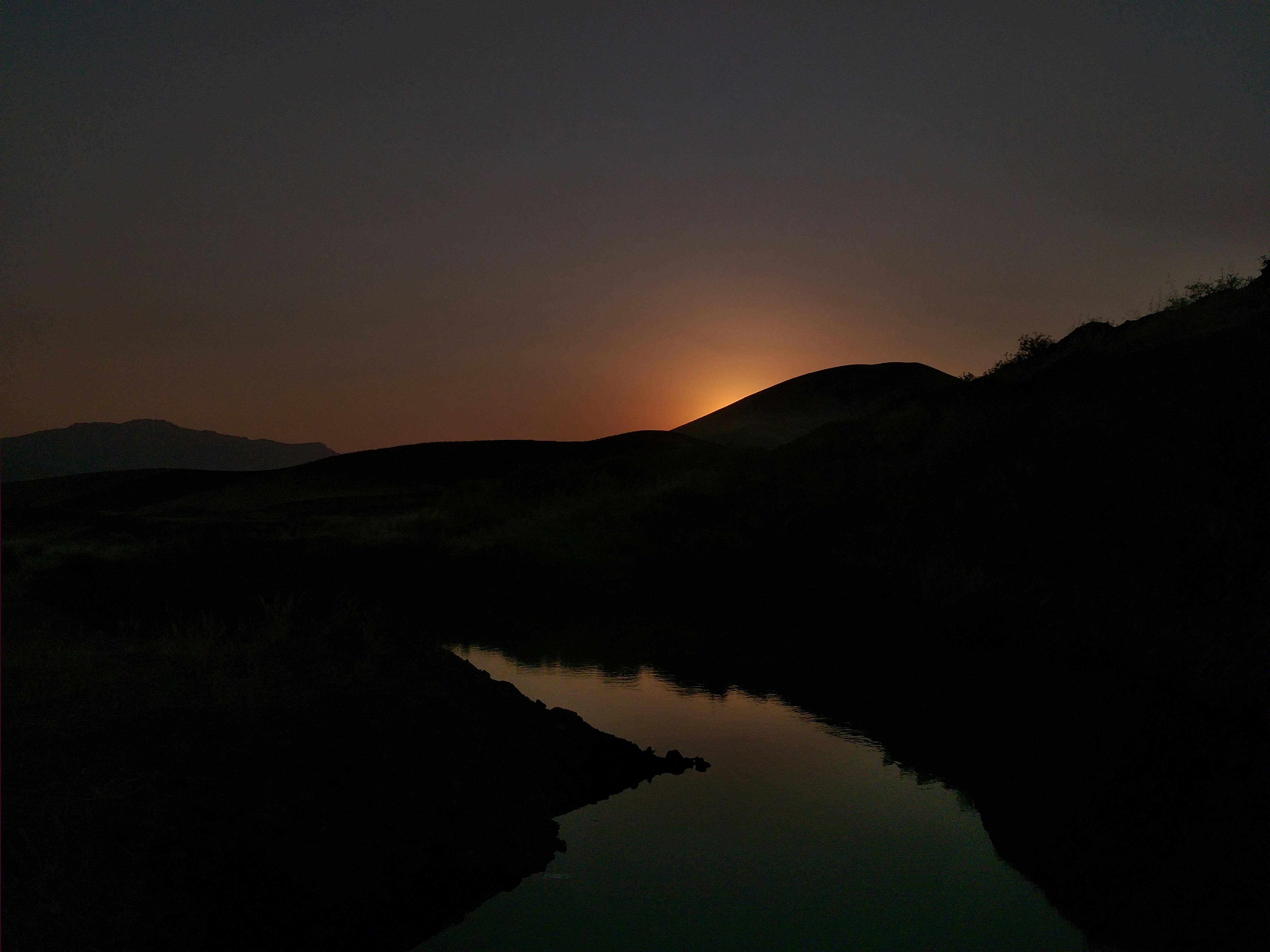 Sunset casts a soft glow over a serene lake surrounded by gentle hills.