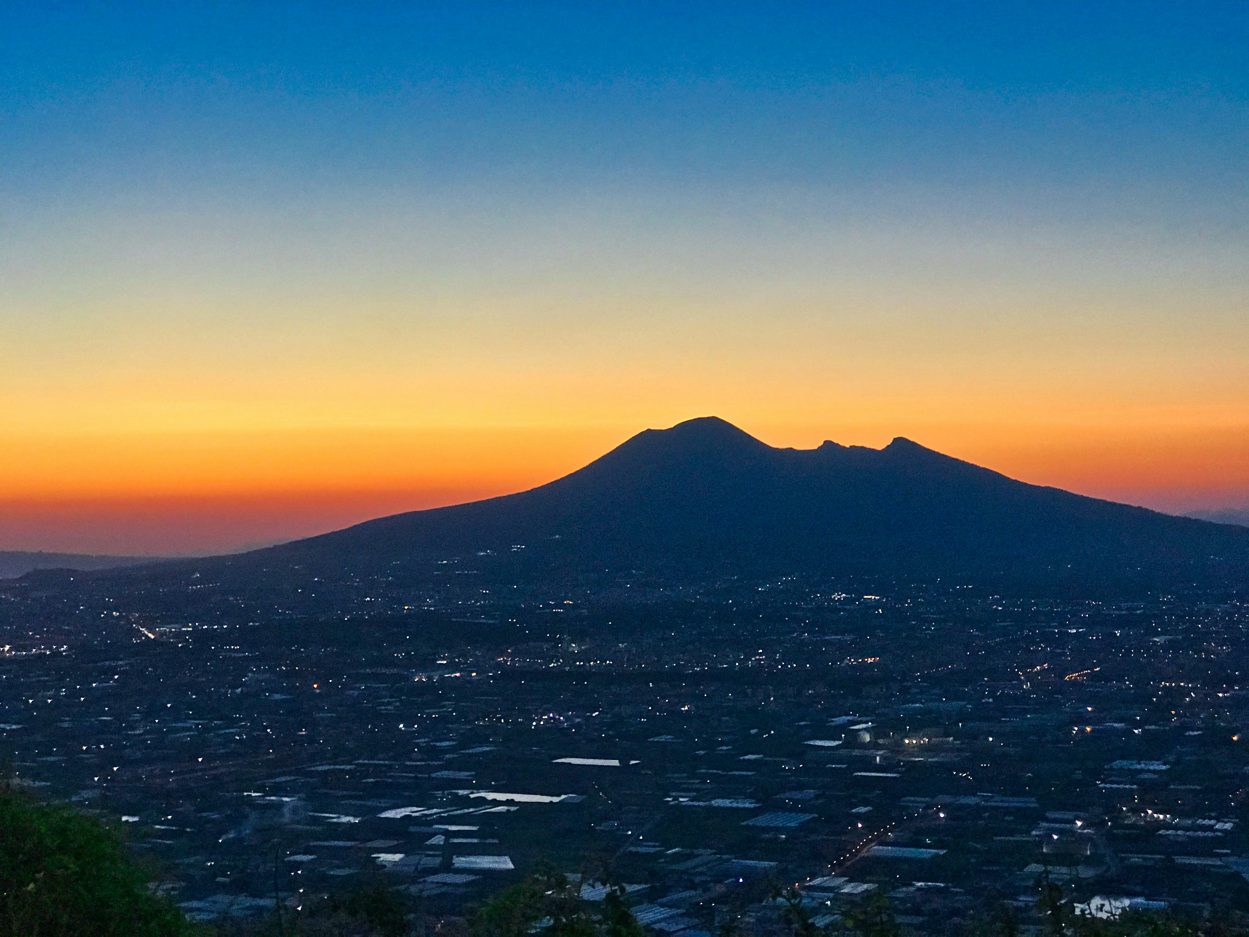 Silhouette of a mountain at sunset with a cityscape below, lights twinkling under a gradient sky.