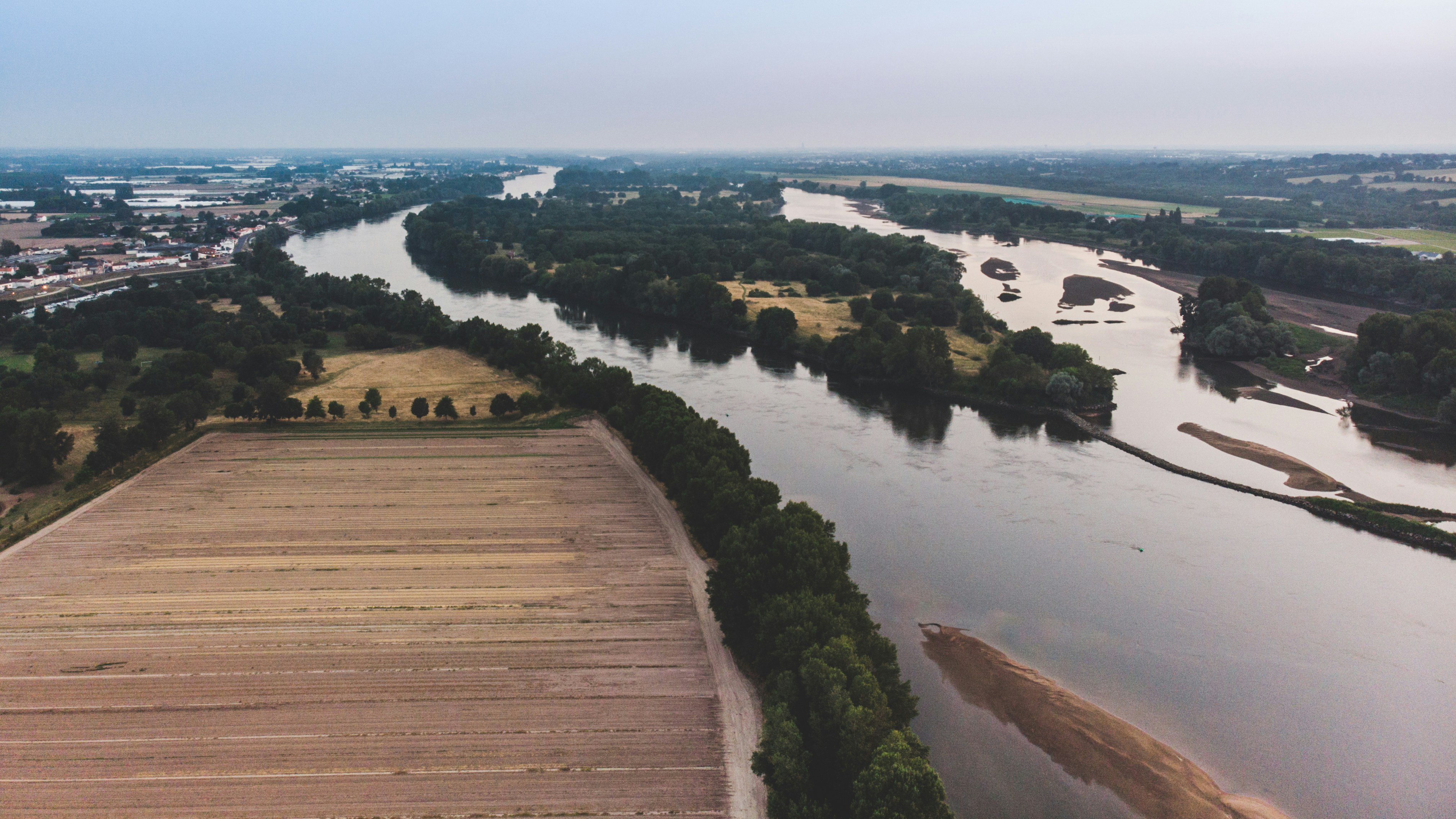 Aerial view of a winding river flanked by lush fields and dense forests under a hazy sky.