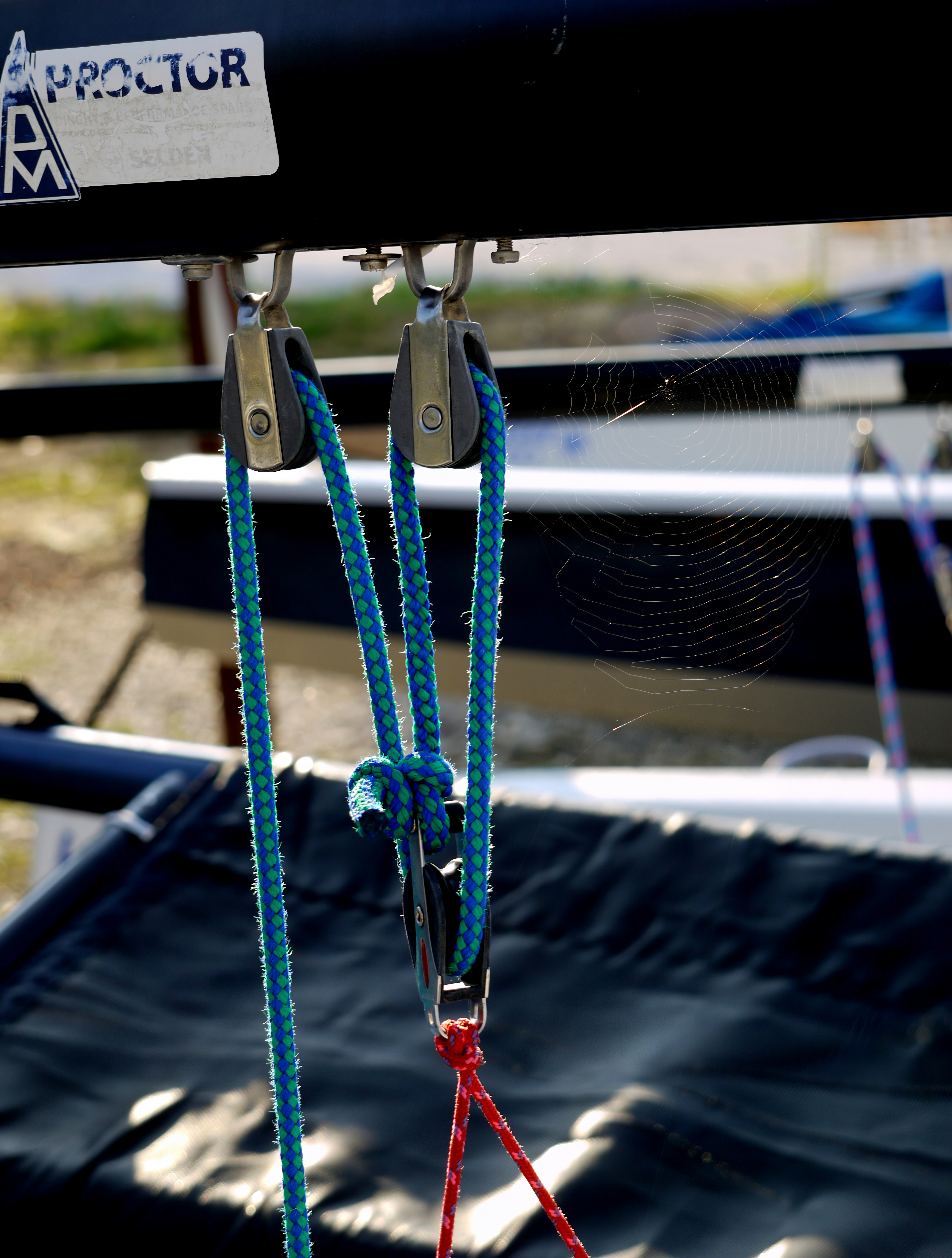 Close-up of turquoise ropes, a red tether, and a metal carabiner on a boat frame, with a delicate spider web catching the light.