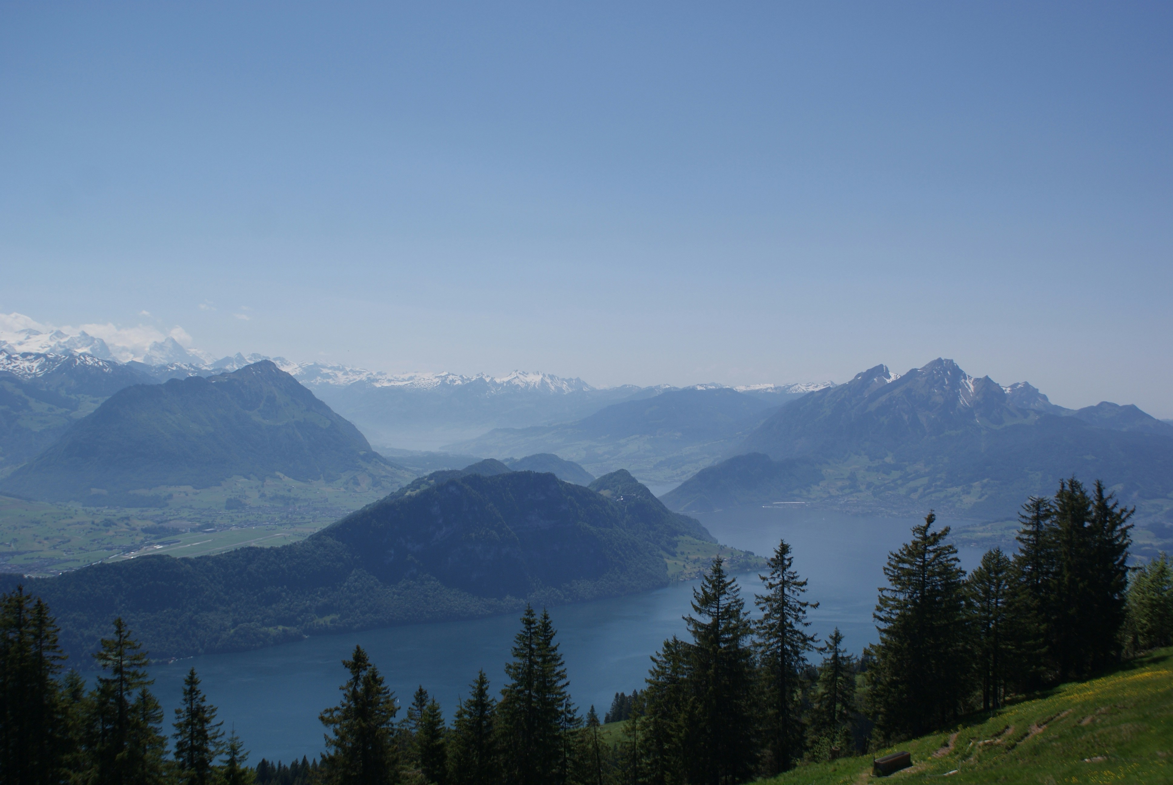 Vast mountain landscape with lush greenery and a tranquil lake, framed by distant snow-capped peaks under a clear blue sky.