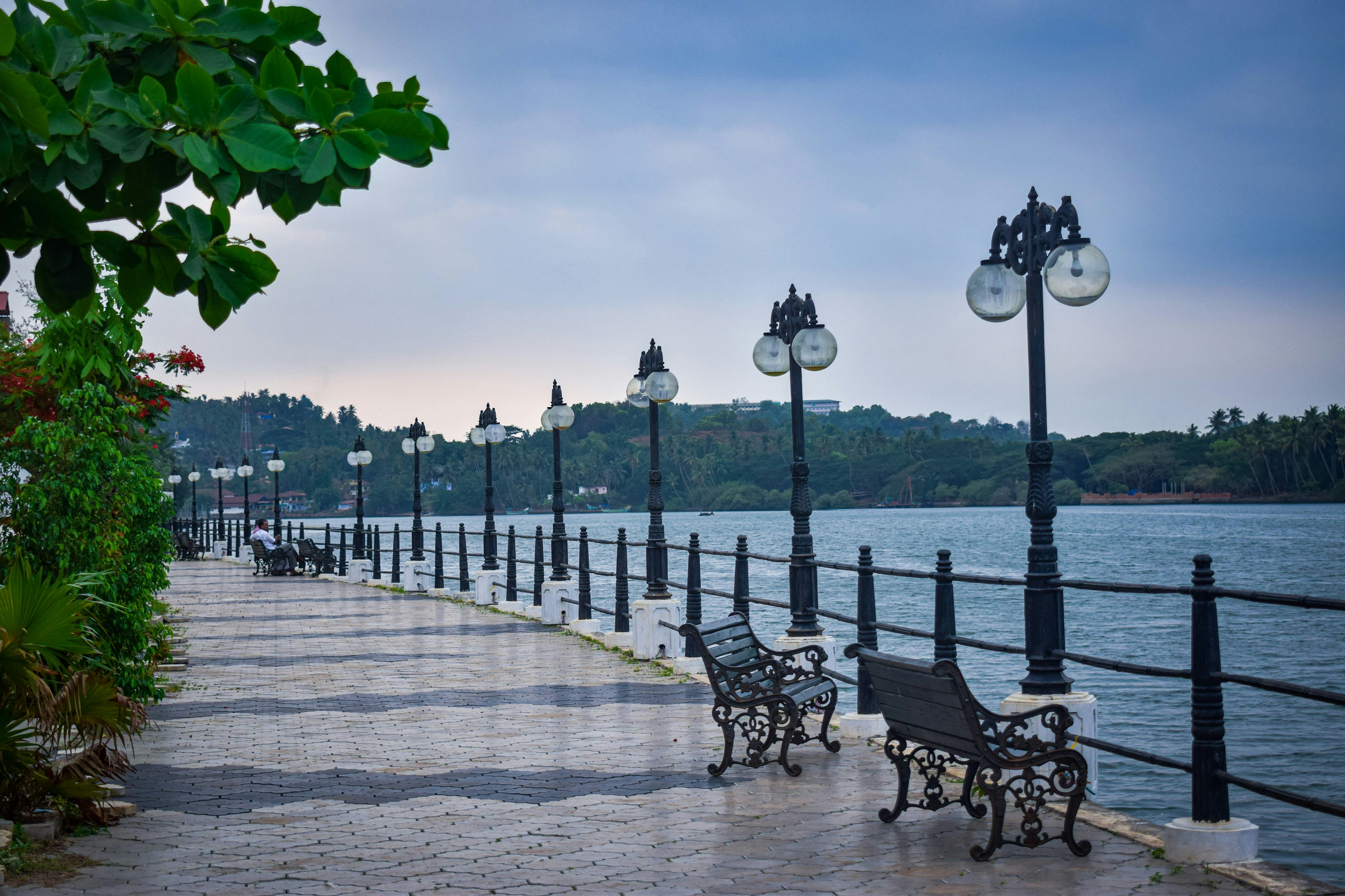 Empty riverside walkway lined with benches and vintage street lamps under a cloudy evening sky.
