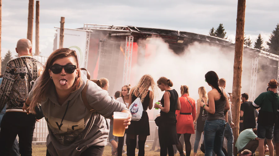Woman in sunglasses leans toward the camera with her tongue out and a drink in hand at an outdoor festival. A smoky stage and clustered attendees fill the background.