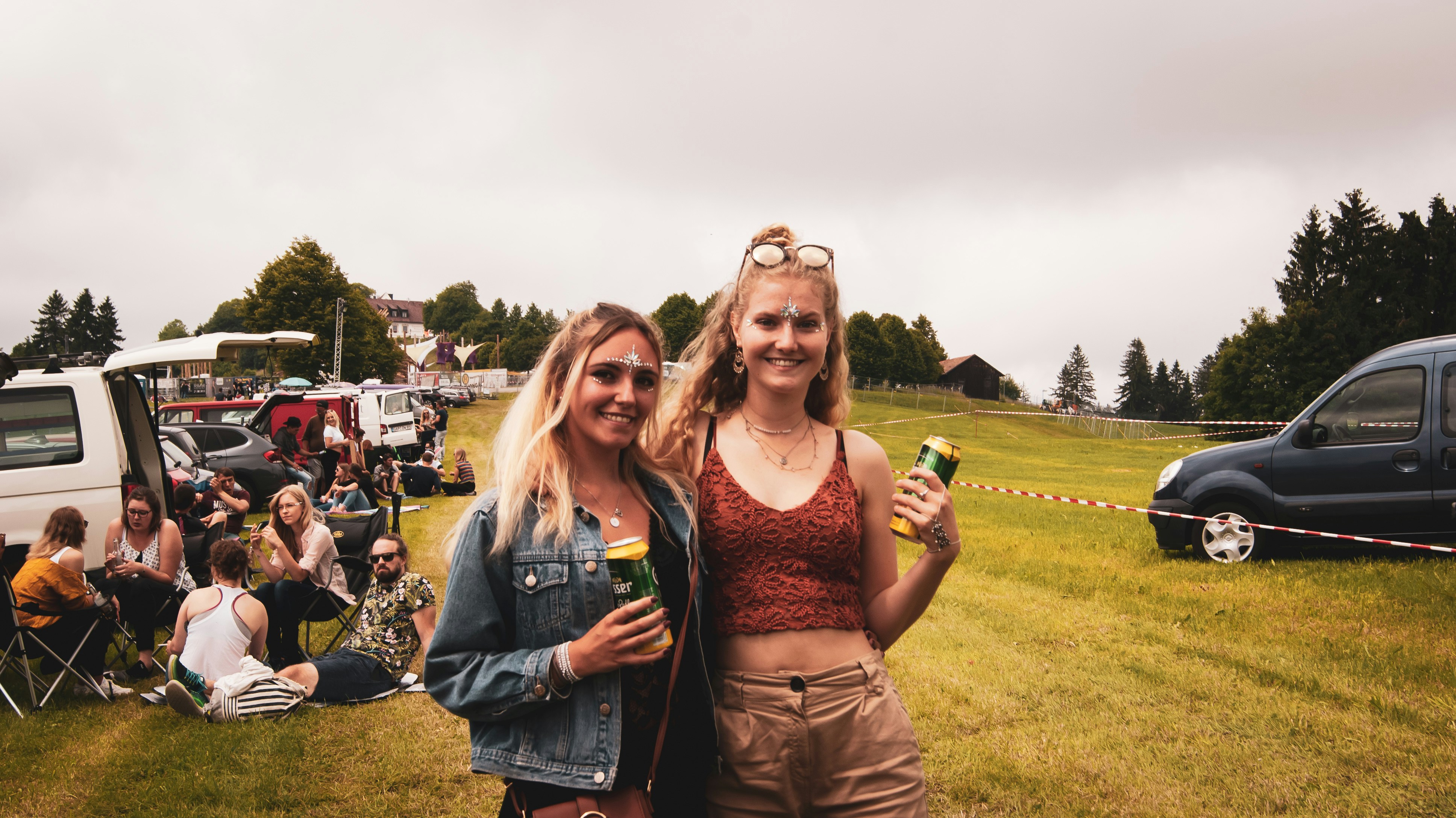 Two women with face paint holding cans, standing in a grassy field with festival-goers and vehicles in the background.