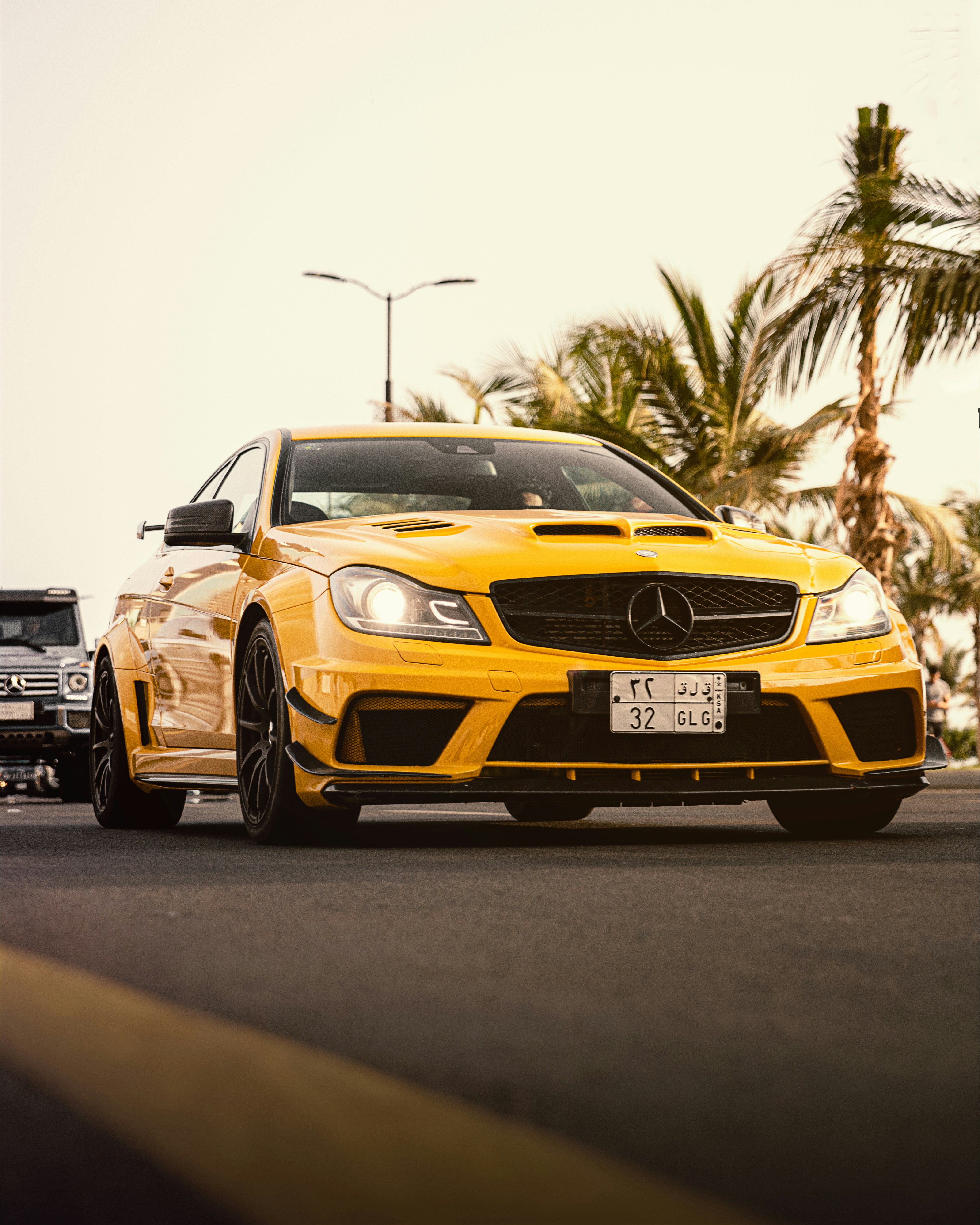 A yellow Mercedes-AMG sedan charges toward the camera on a sunlit street, its aggressive front grille and headlights in clear detail. Palm trees silhouette the background as a second vehicle trails behind.