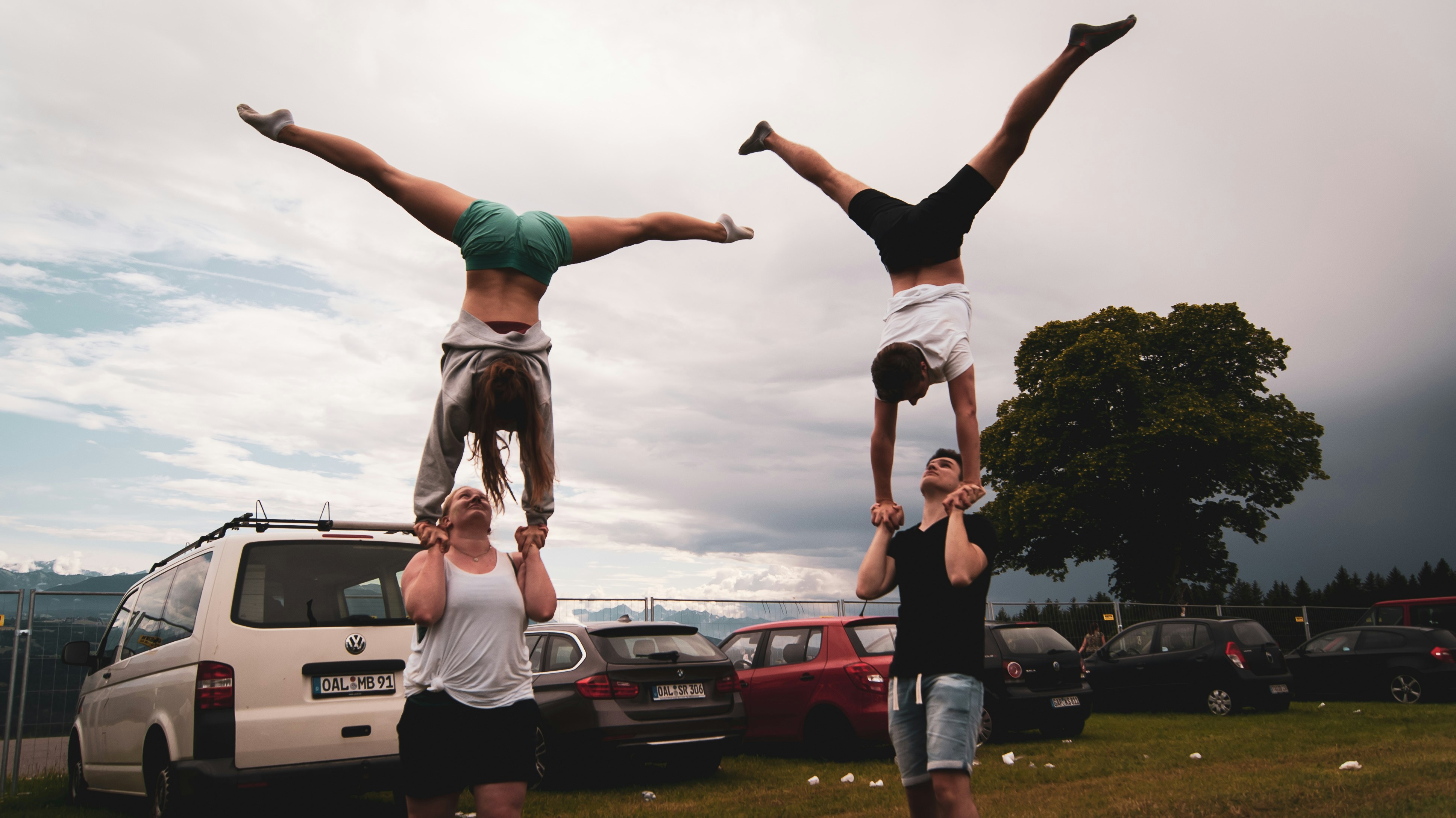Two friends perform handstands overhead in a cluttered parking lot, with cars and a tree in the background, capturing a spontaneous athletic moment.