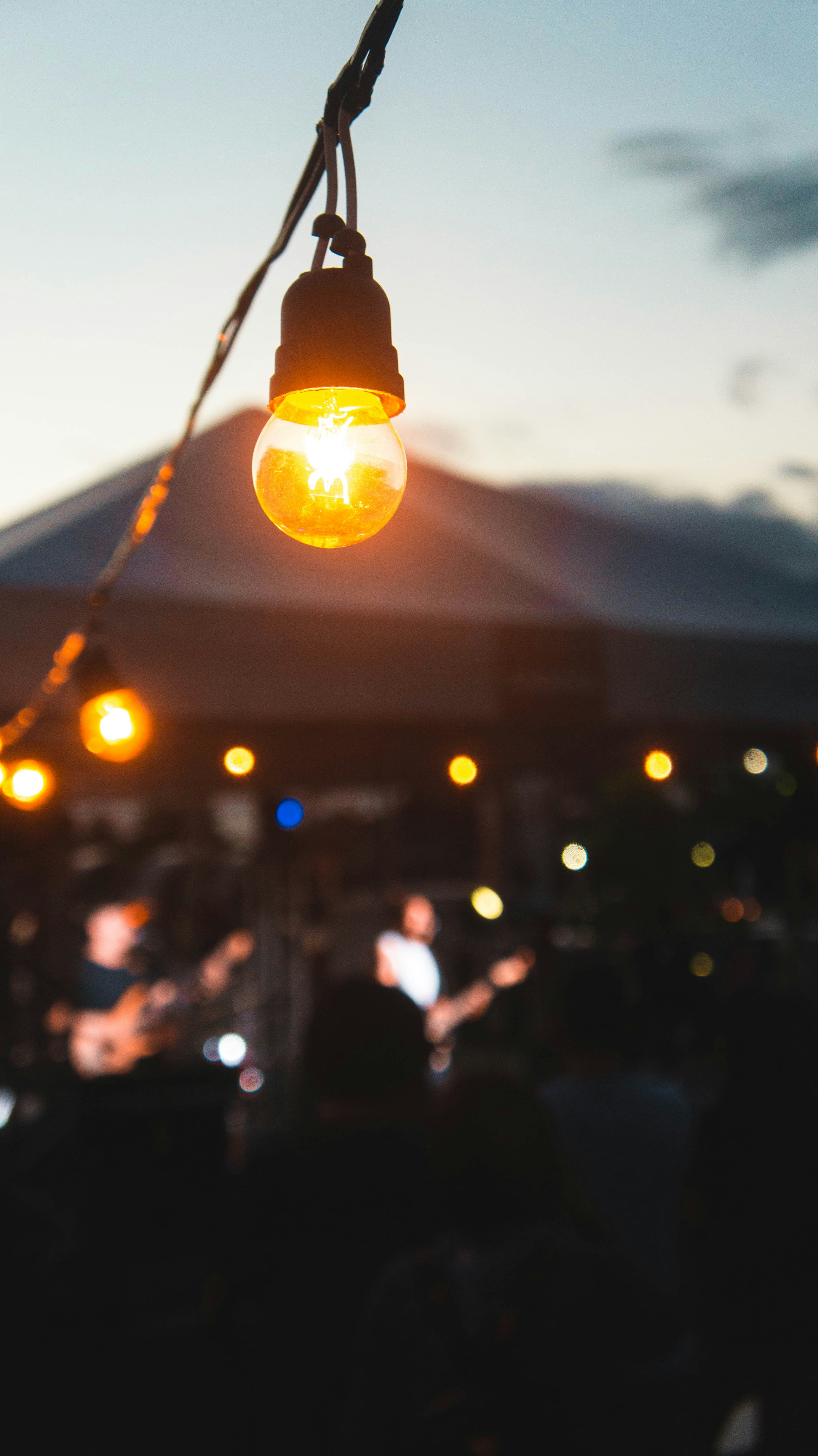 A glowing light bulb hangs in the foreground, illuminating a blurred scene of musicians performing on stage at dusk.