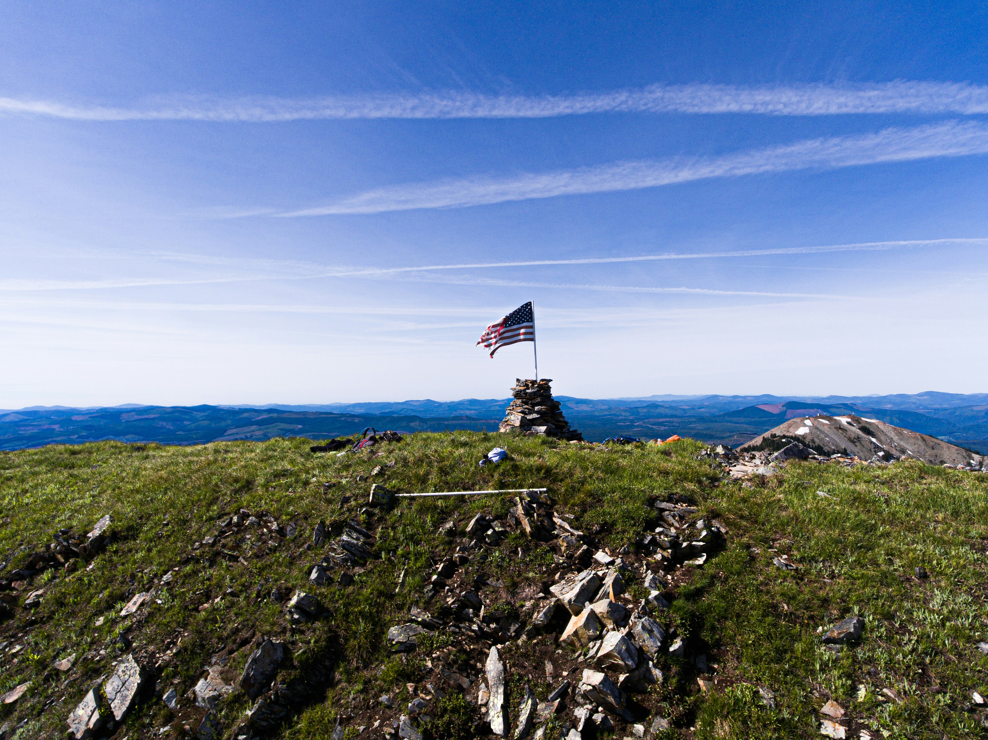 American flag waving atop a rocky summit, surrounded by lush greenery and distant mountains under a clear blue sky.
