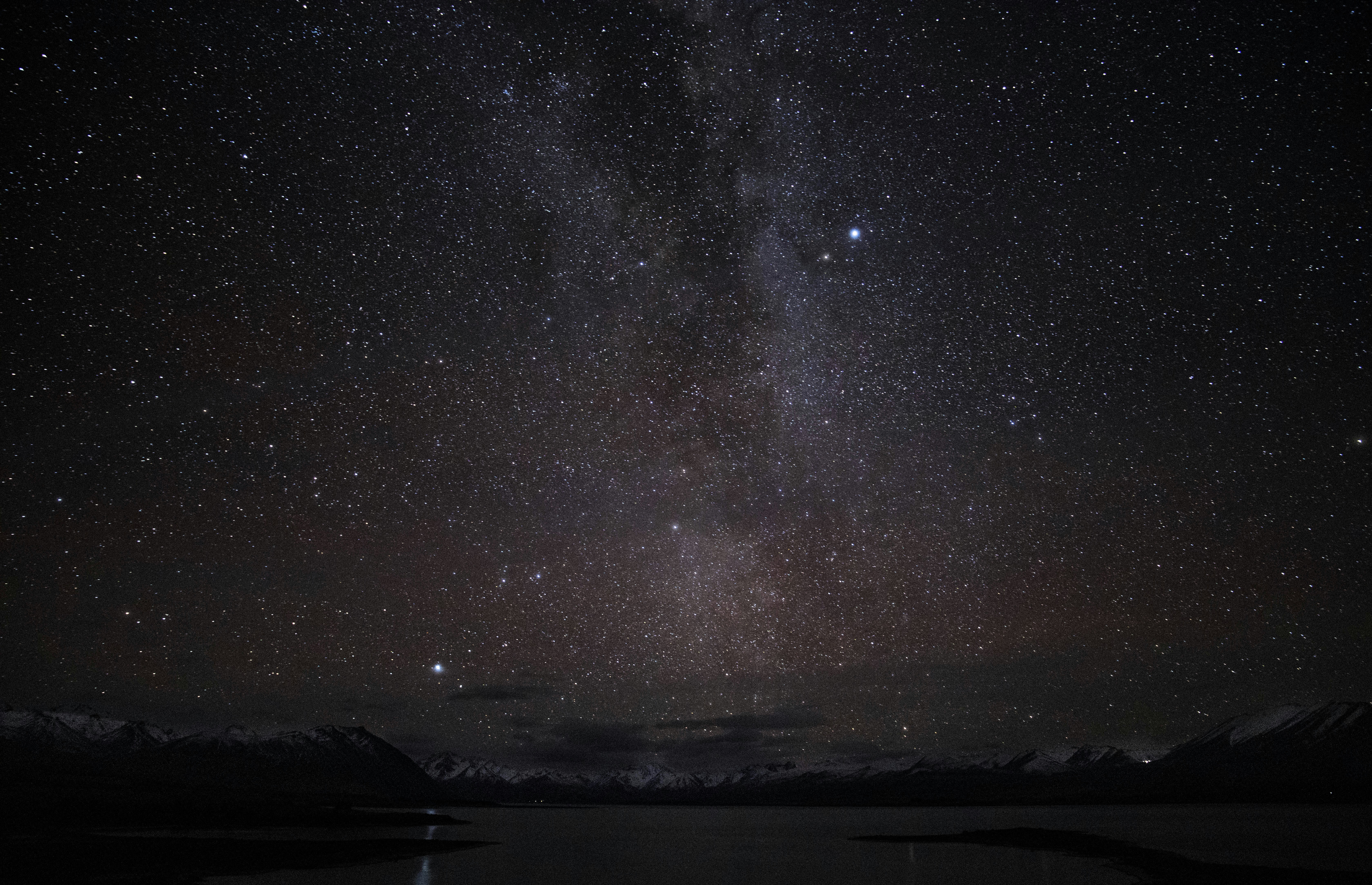 Starry night sky above silhouetted mountain range and calm lake.