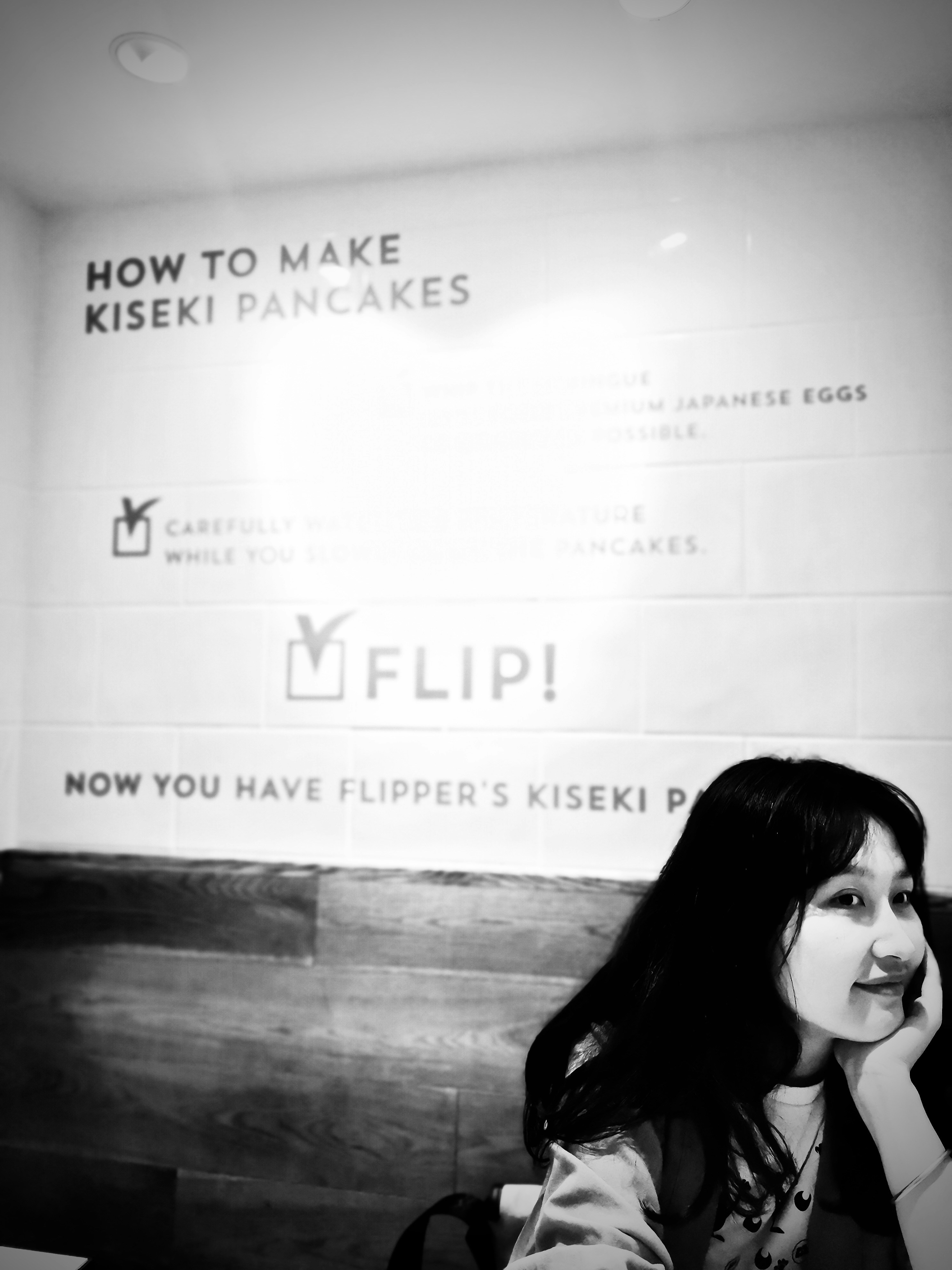Candid portrait of a person seated against a wall with Kiseki Pancakes signage, captured in black and white.