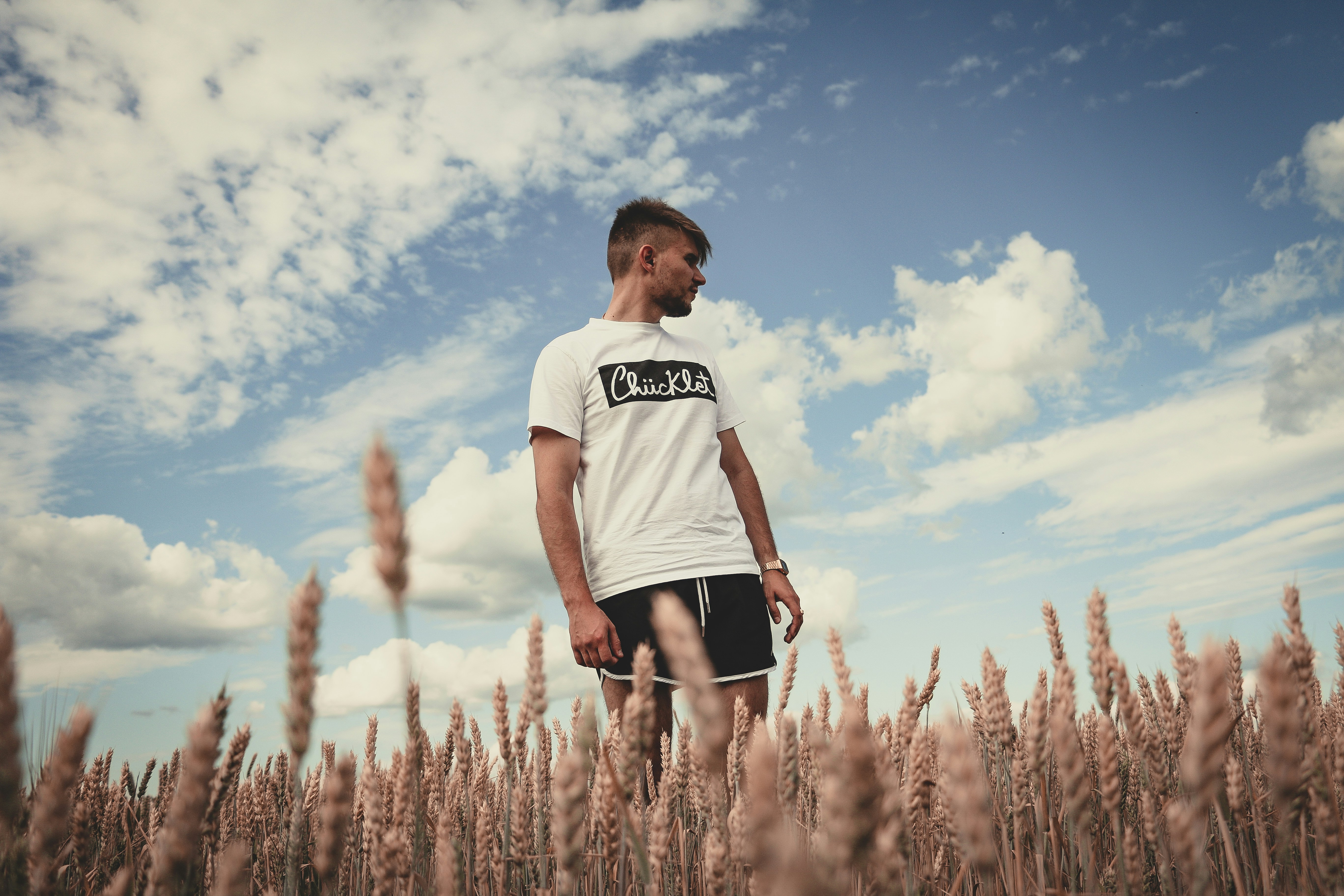 Young man standing amidst golden wheat under a vast sky dotted with clouds.