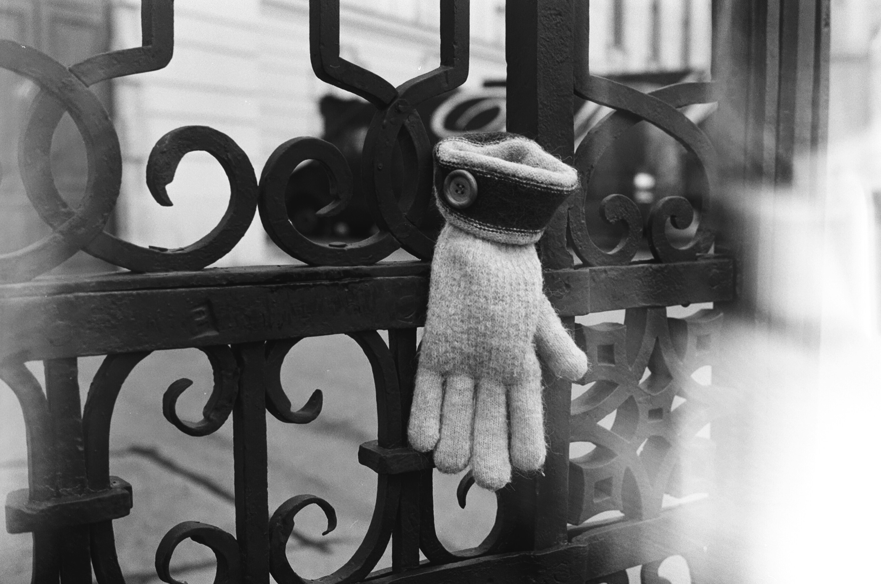 Gloved hand rests on an intricate wrought-iron fence, creating a solitary focal point in a monochrome scene. A quiet, urban moment highlighted by soft blur and high contrast.