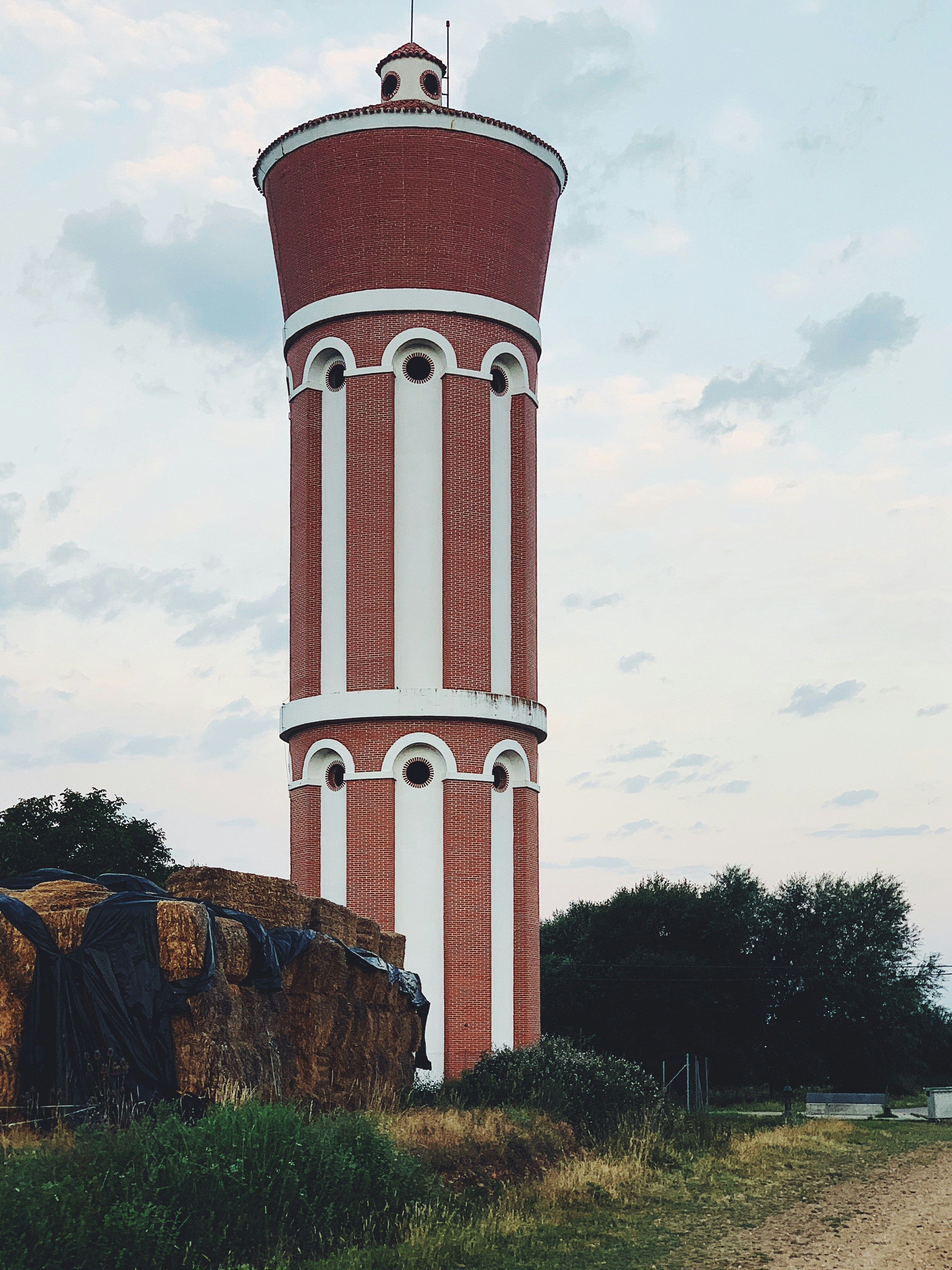 A distinctive water tower with red and white stripes stands tall against a cloudy sky, surrounded by hay bales and greenery.
