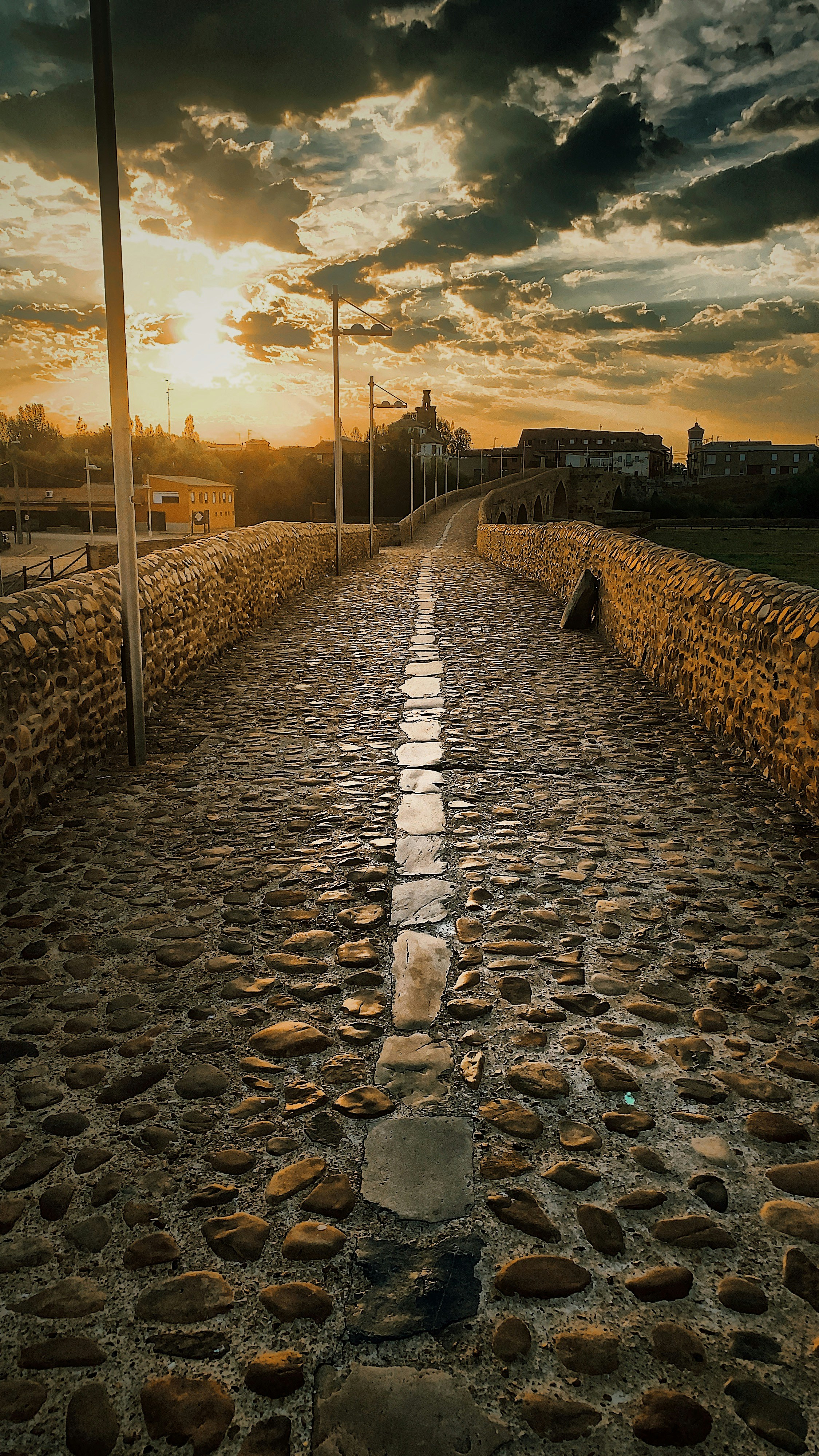 Cobblestone pathway leading towards a sunset, flanked by street lamps and historical buildings in the distance.