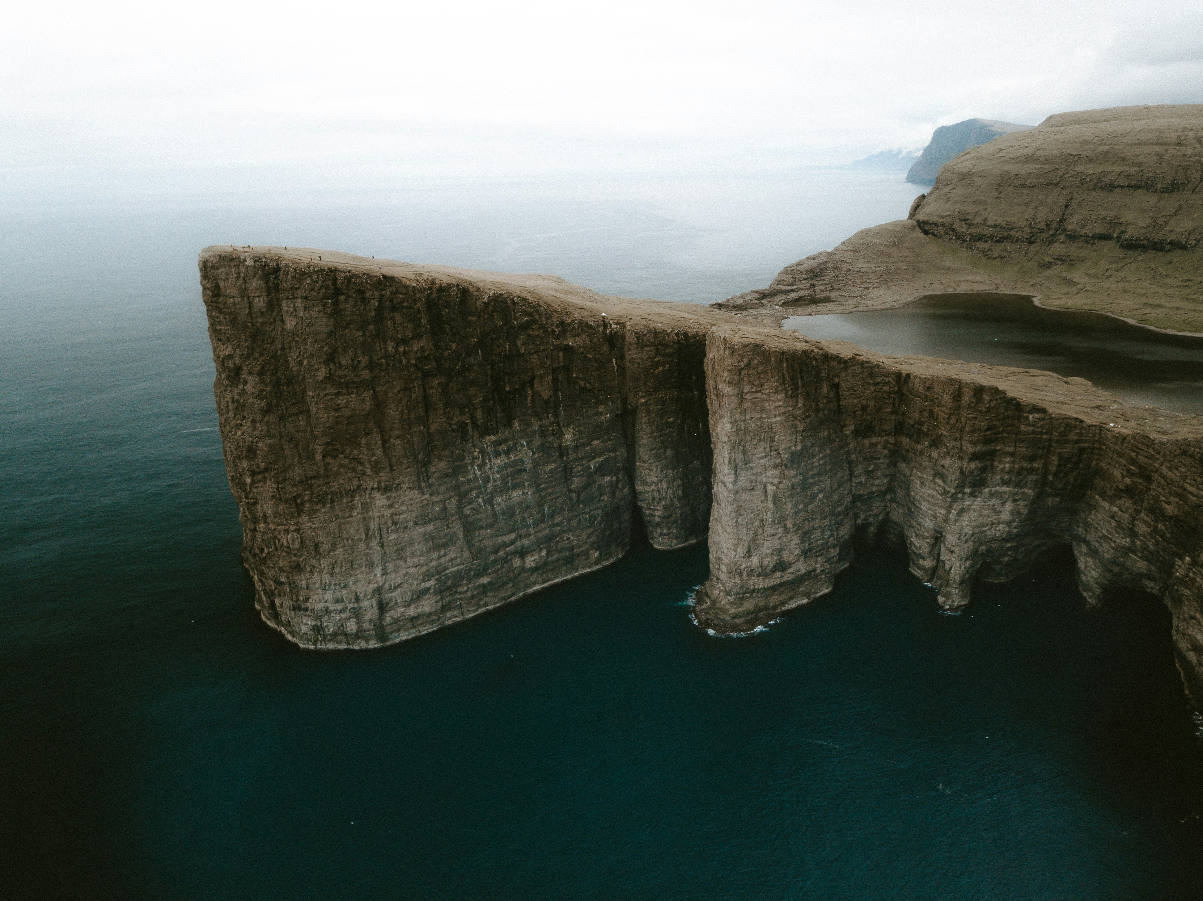 Dramatic cliff jutting into the ocean with a serene lake atop, surrounded by misty skies.