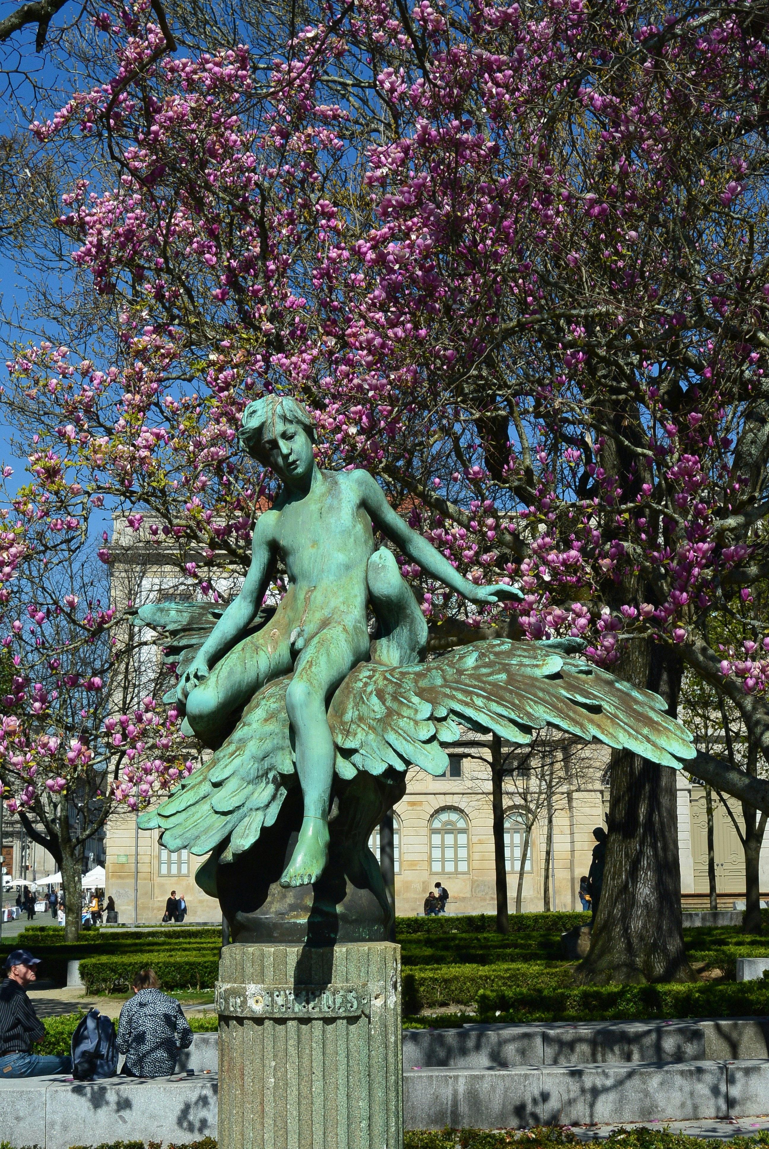 Bronze patina statue of a winged child perched on a pedestal, framed by pink blossoms in a sunlit city park.