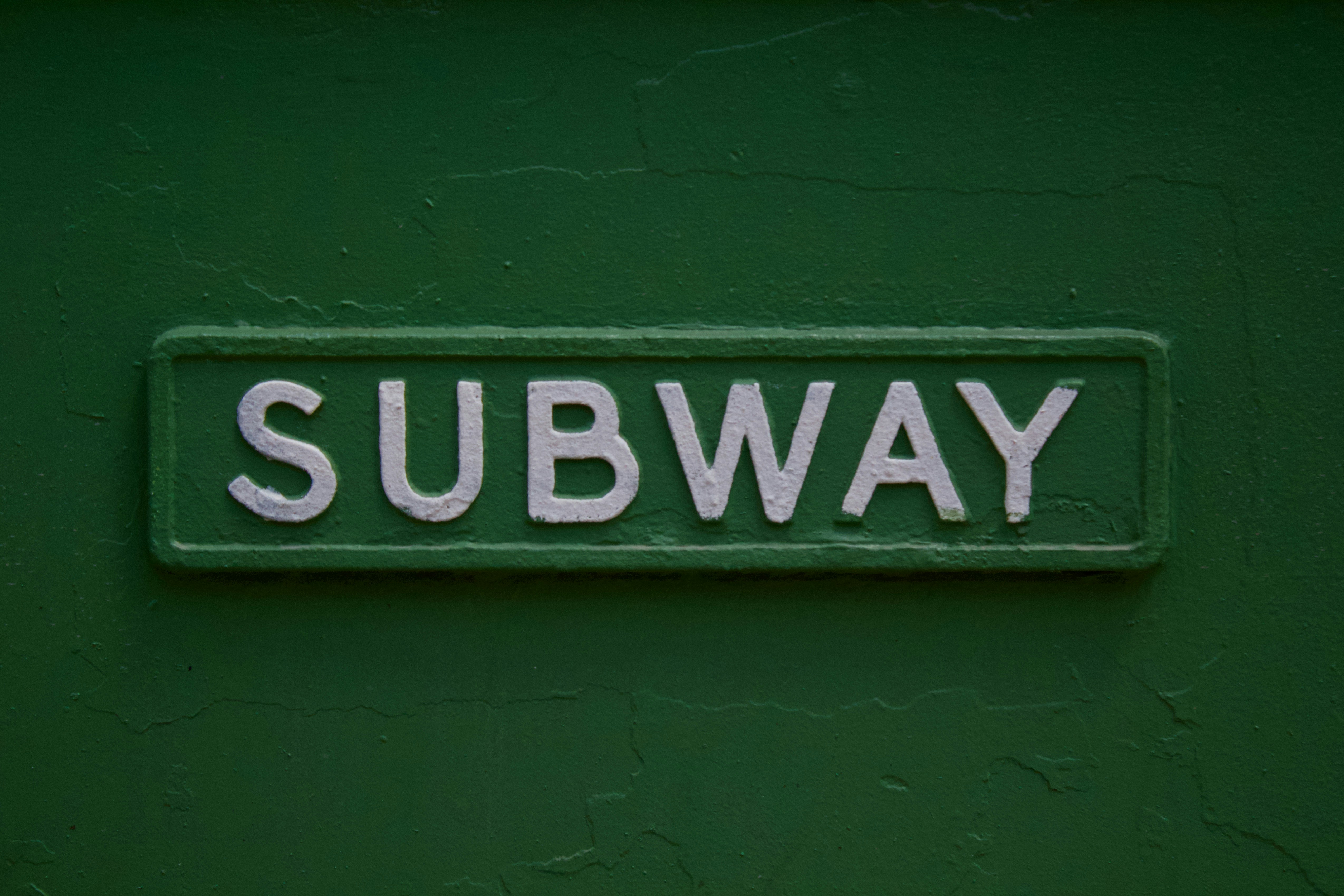 Green subway sign with white lettering, prominently displaying 'SUBWAY'. The sign's texture and color contrast highlight its urban significance.