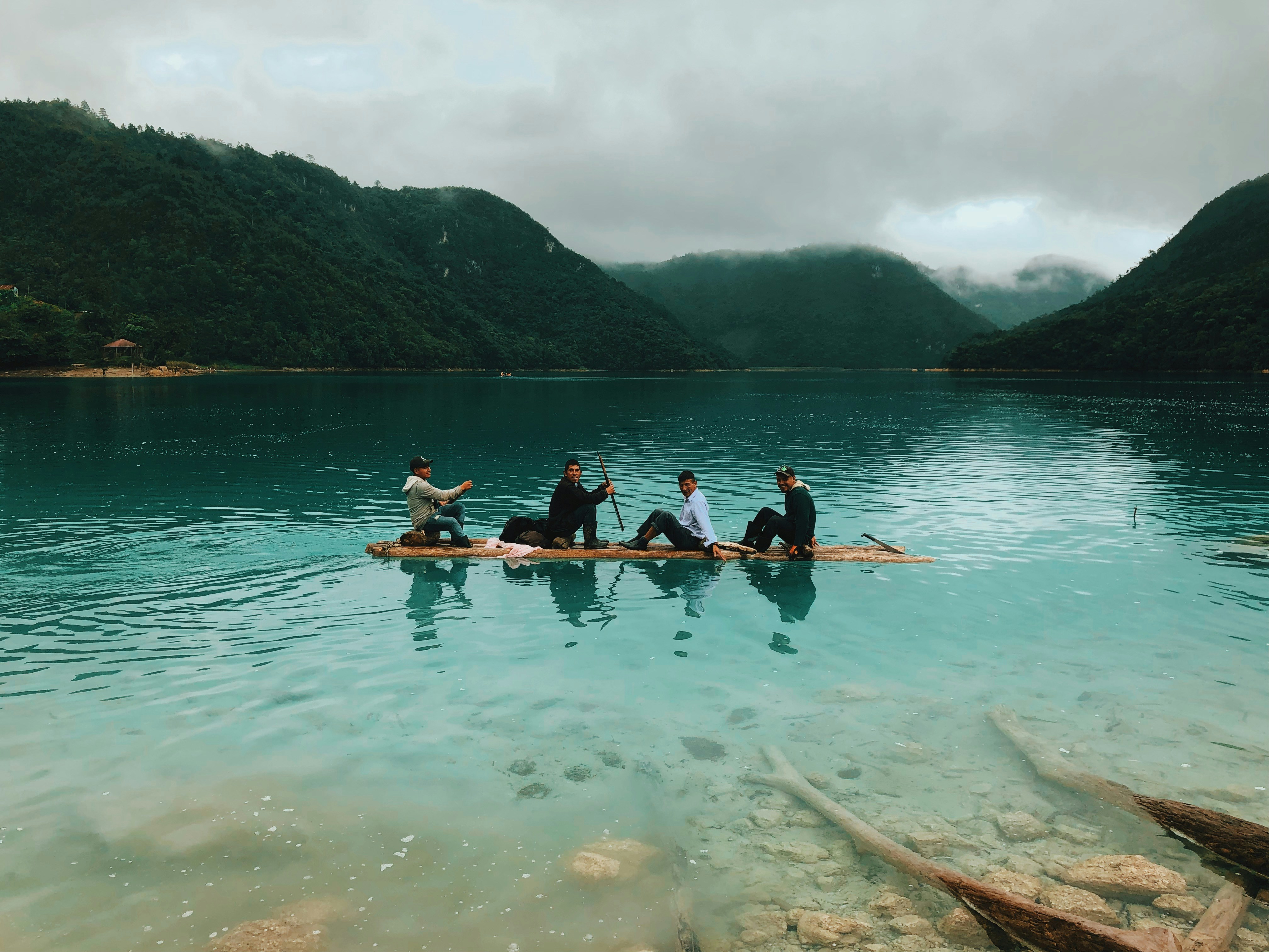 Four individuals navigating a tranquil lake on a bamboo raft, surrounded by lush green mountains and a cloudy sky.