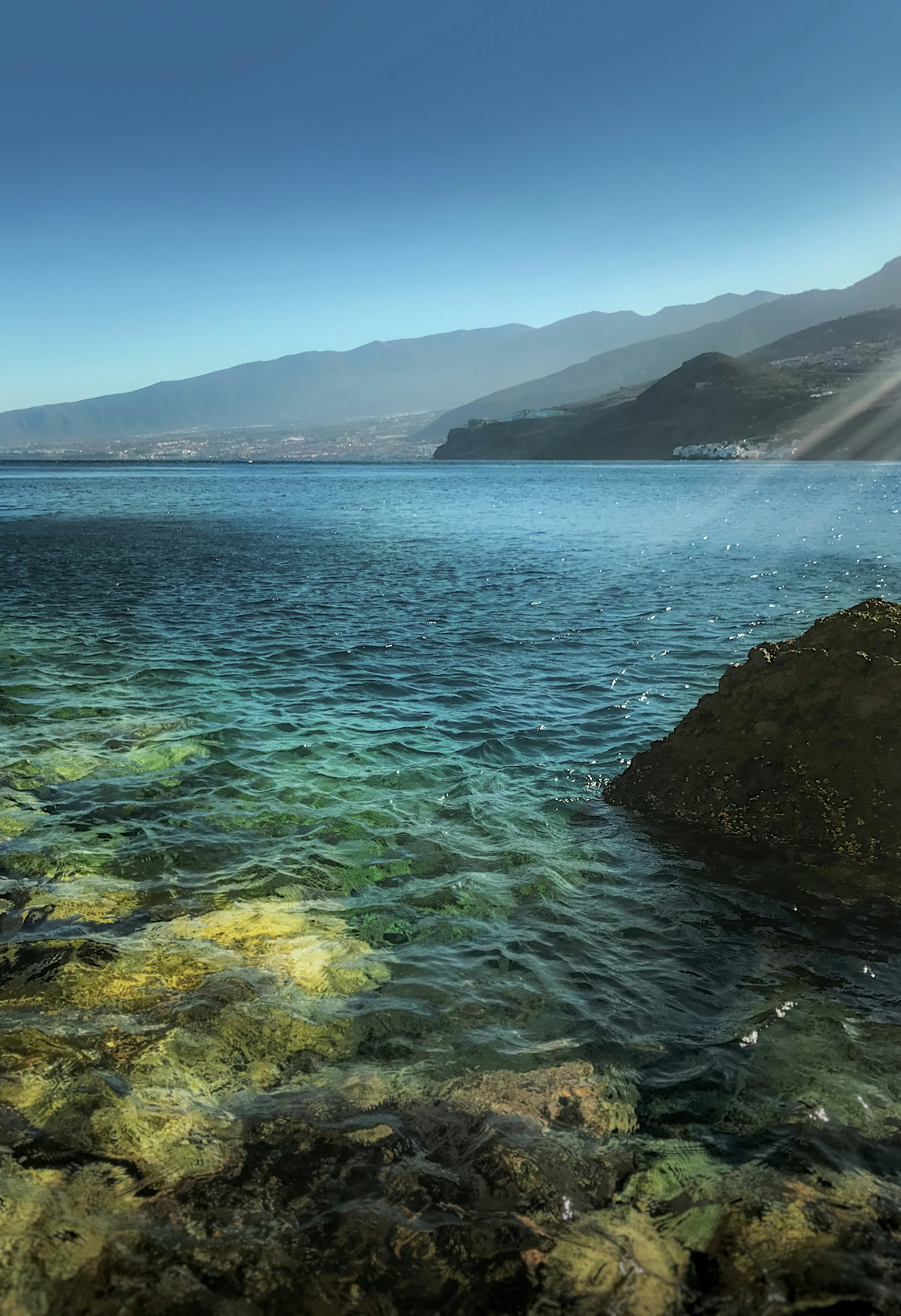 Clear turquoise waters gently lap against rocky shore, with distant mountains framing the horizon. The scene evokes a sense of tranquility and natural beauty.