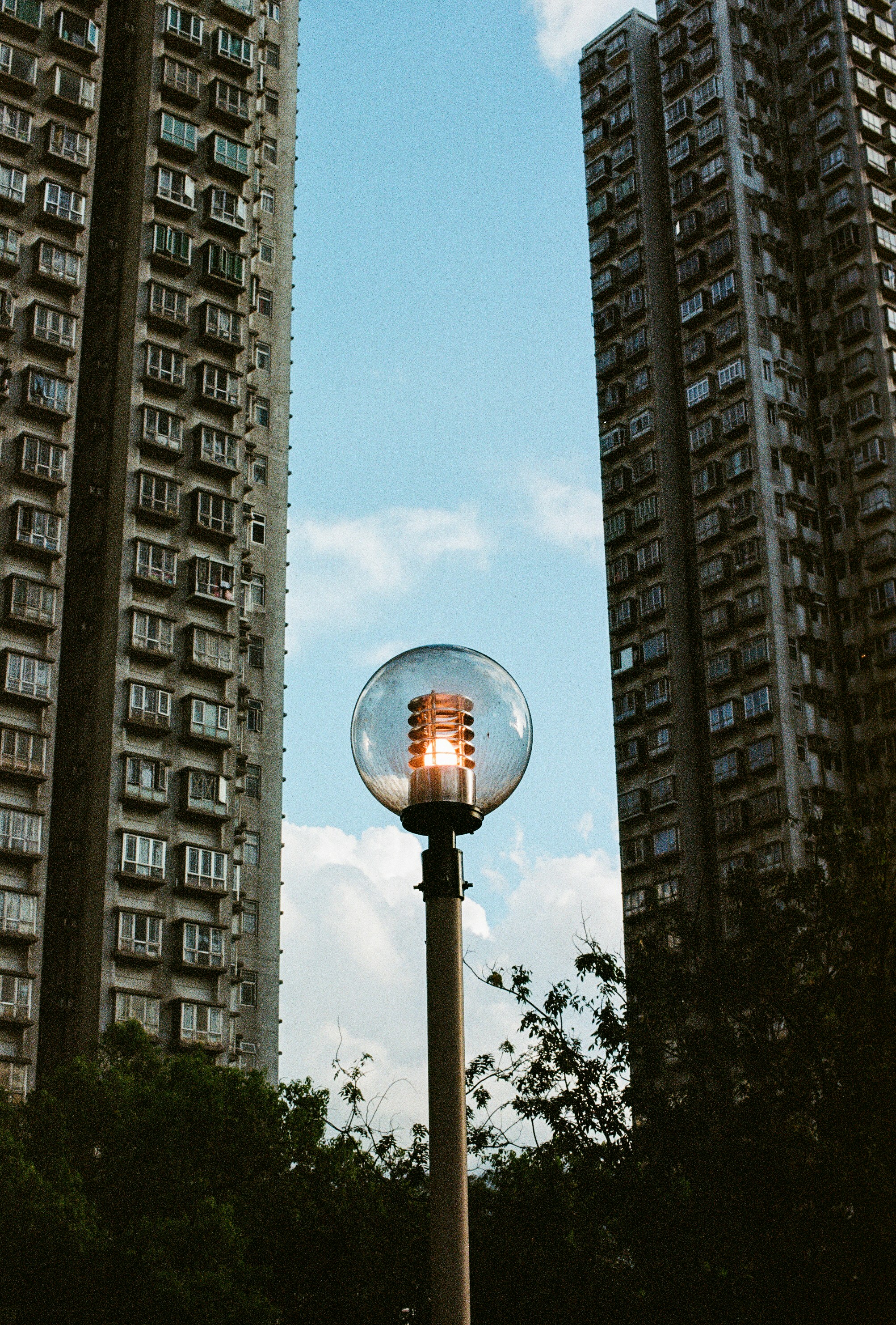 A glowing street lamp stands tall between towering residential buildings, highlighting the contrast of urban life against a bright sky.
