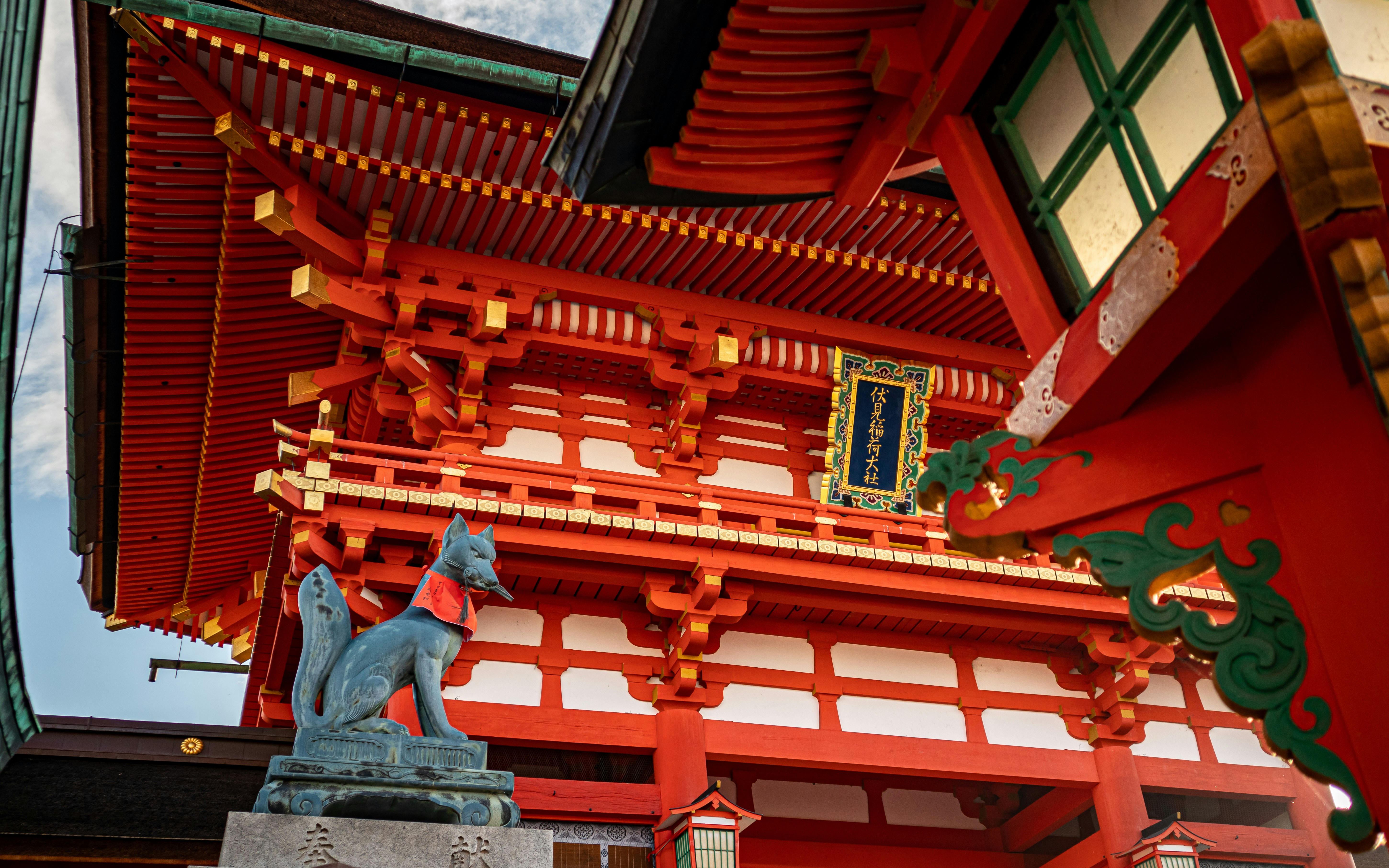A stone fox statue adorned with a red scarf stands proudly in front of a vibrant shrine, showcasing intricate architectural details and rich colors.
