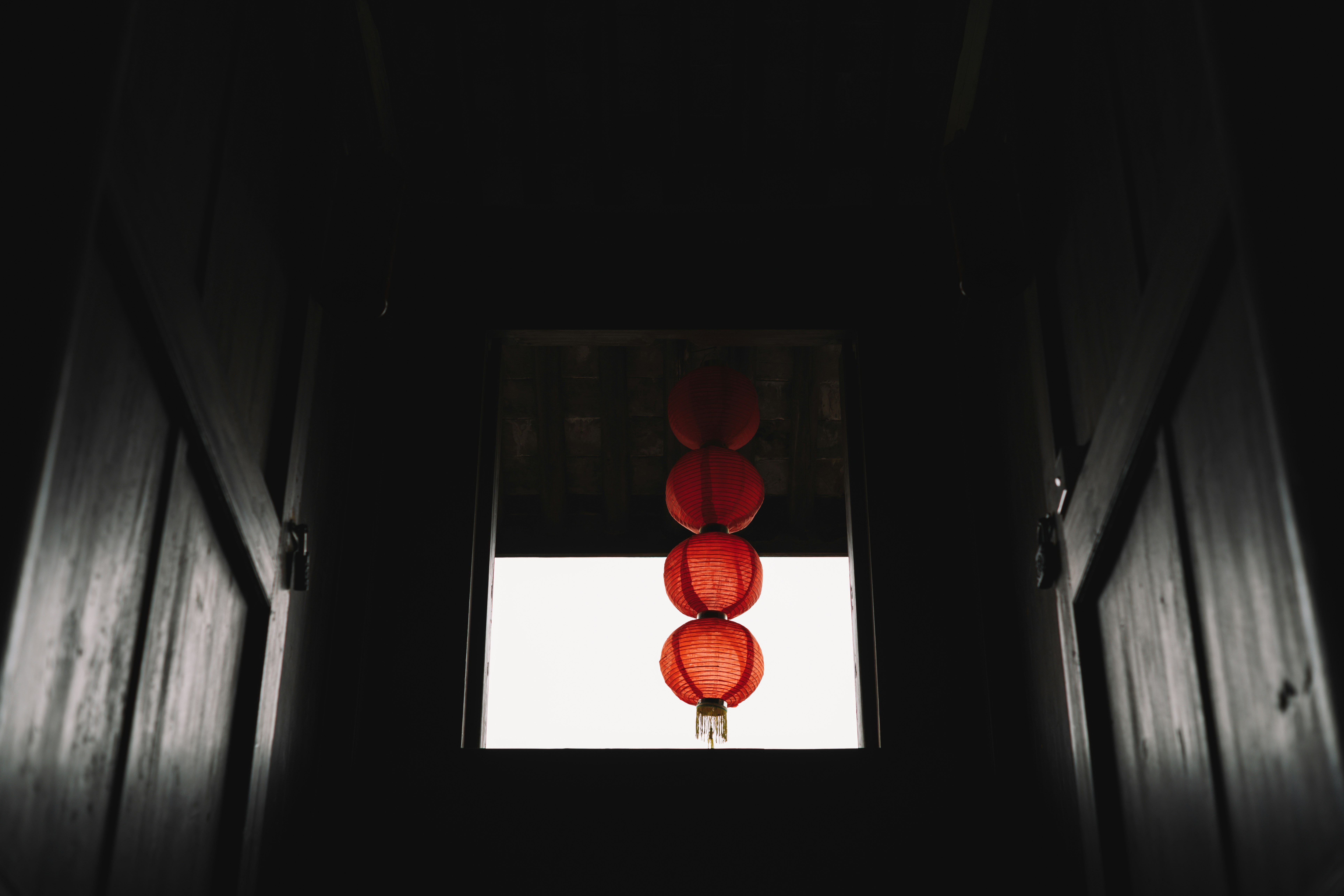 Three vibrant red lanterns hang against a bright backdrop, framed by dark wooden doors. The composition evokes a sense of cultural warmth and inviting mystery.