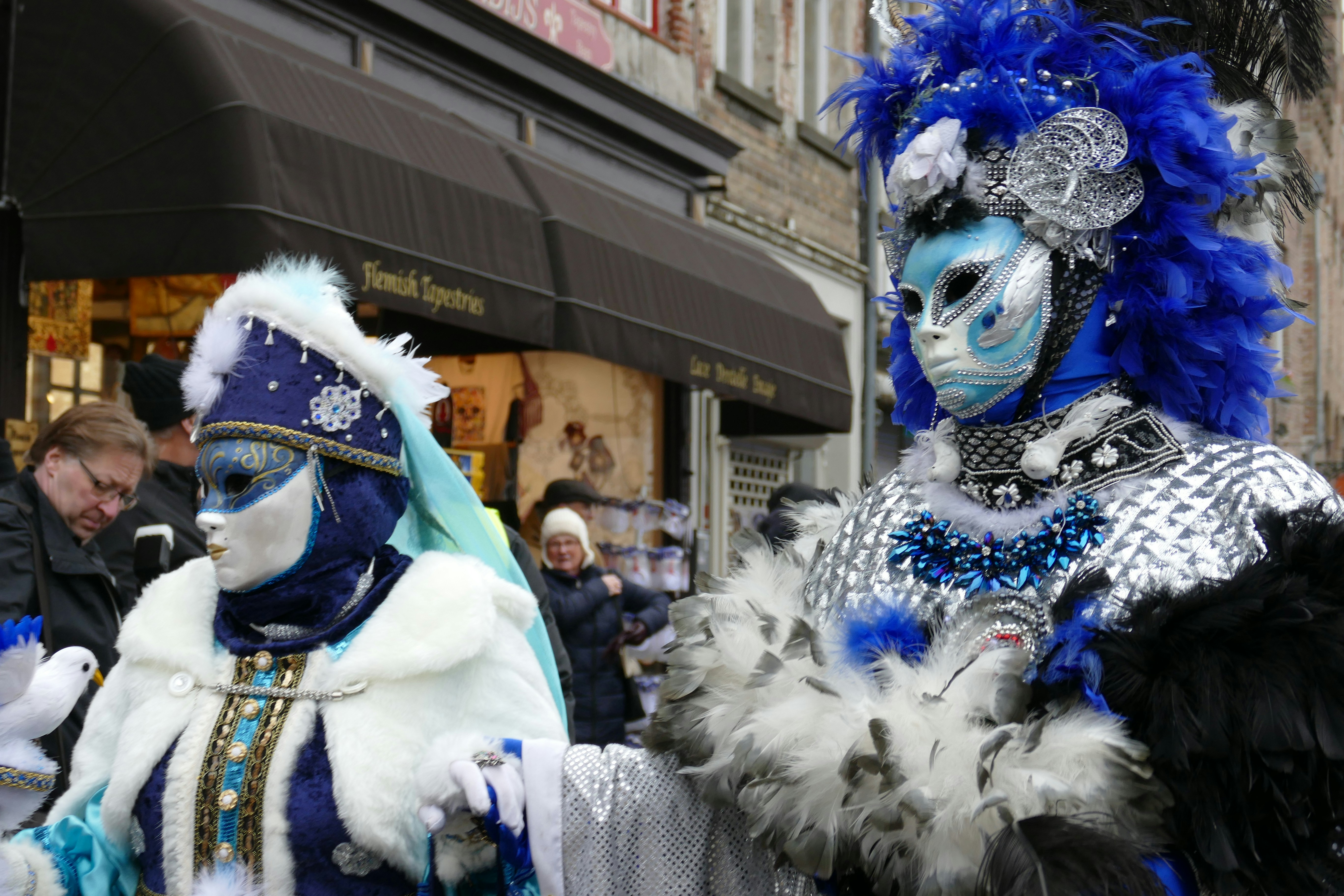 Colorful masked figures in ornate costumes adorned with feathers and jewels during a street procession.