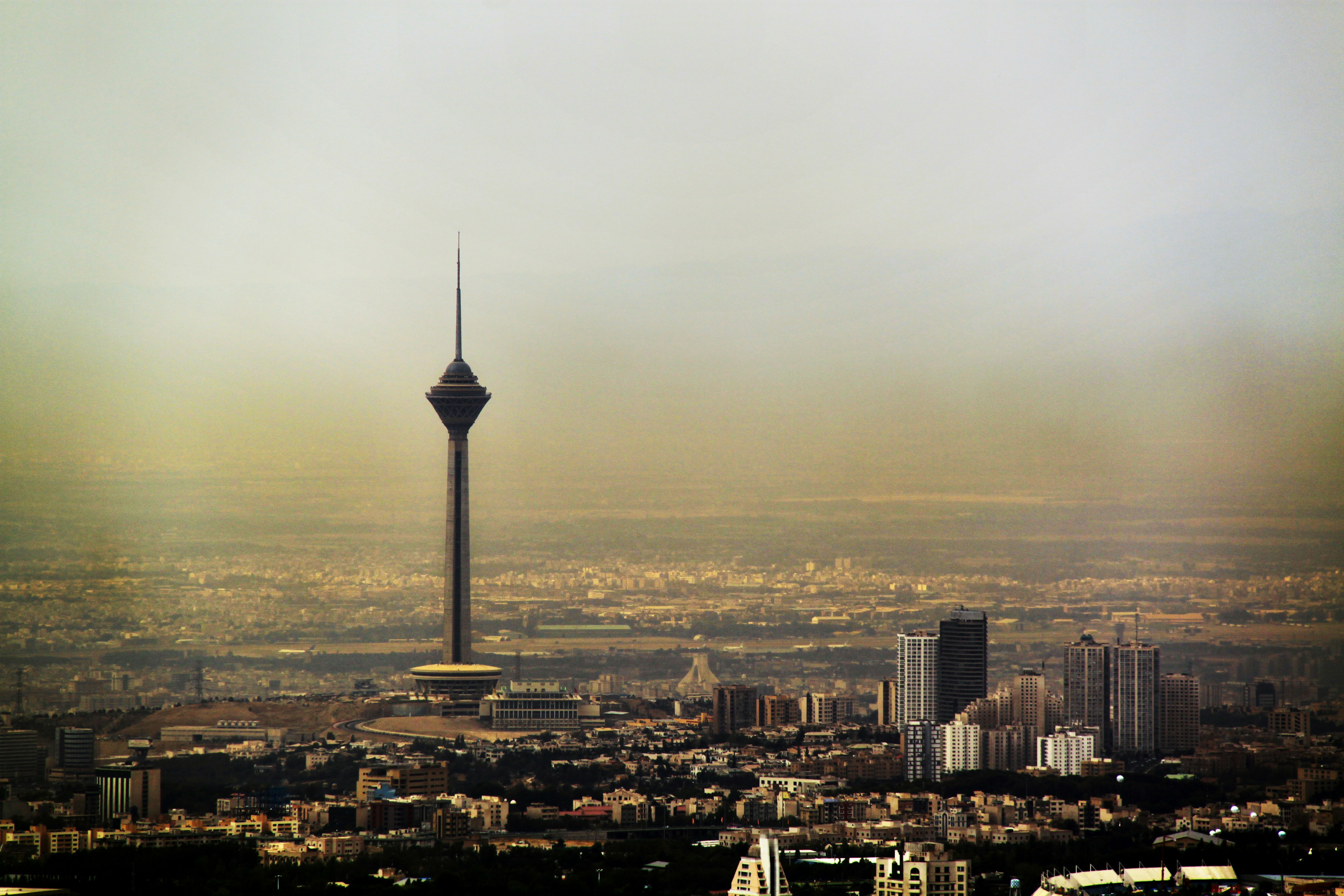 Milad Tower rises majestically over Tehran, surrounded by a sprawling urban landscape under a hazy sky.