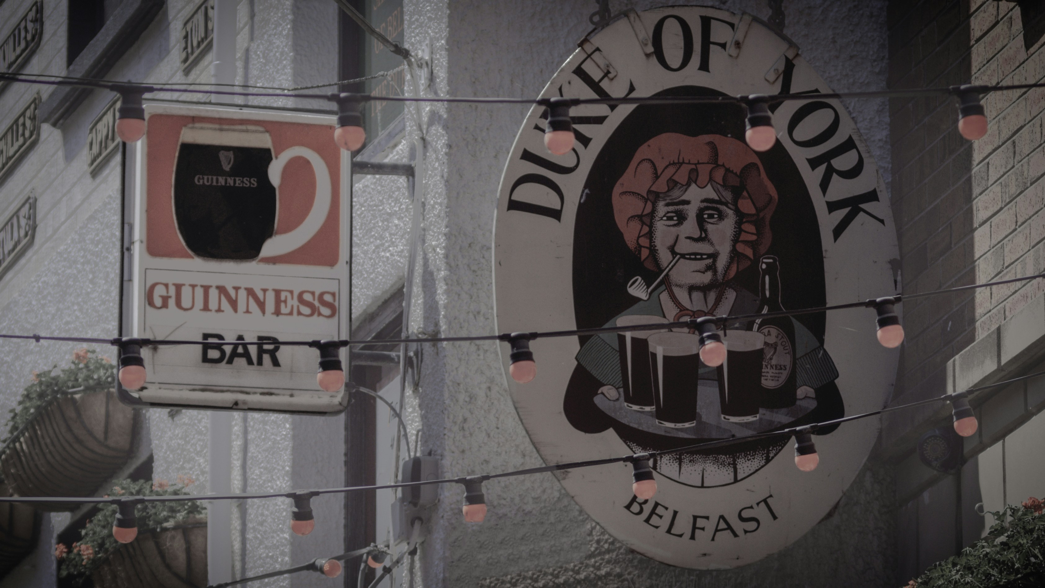 Vintage pub signs for Duke of York and Guinness illuminated by string lights, capturing the essence of Belfast's lively bar scene.