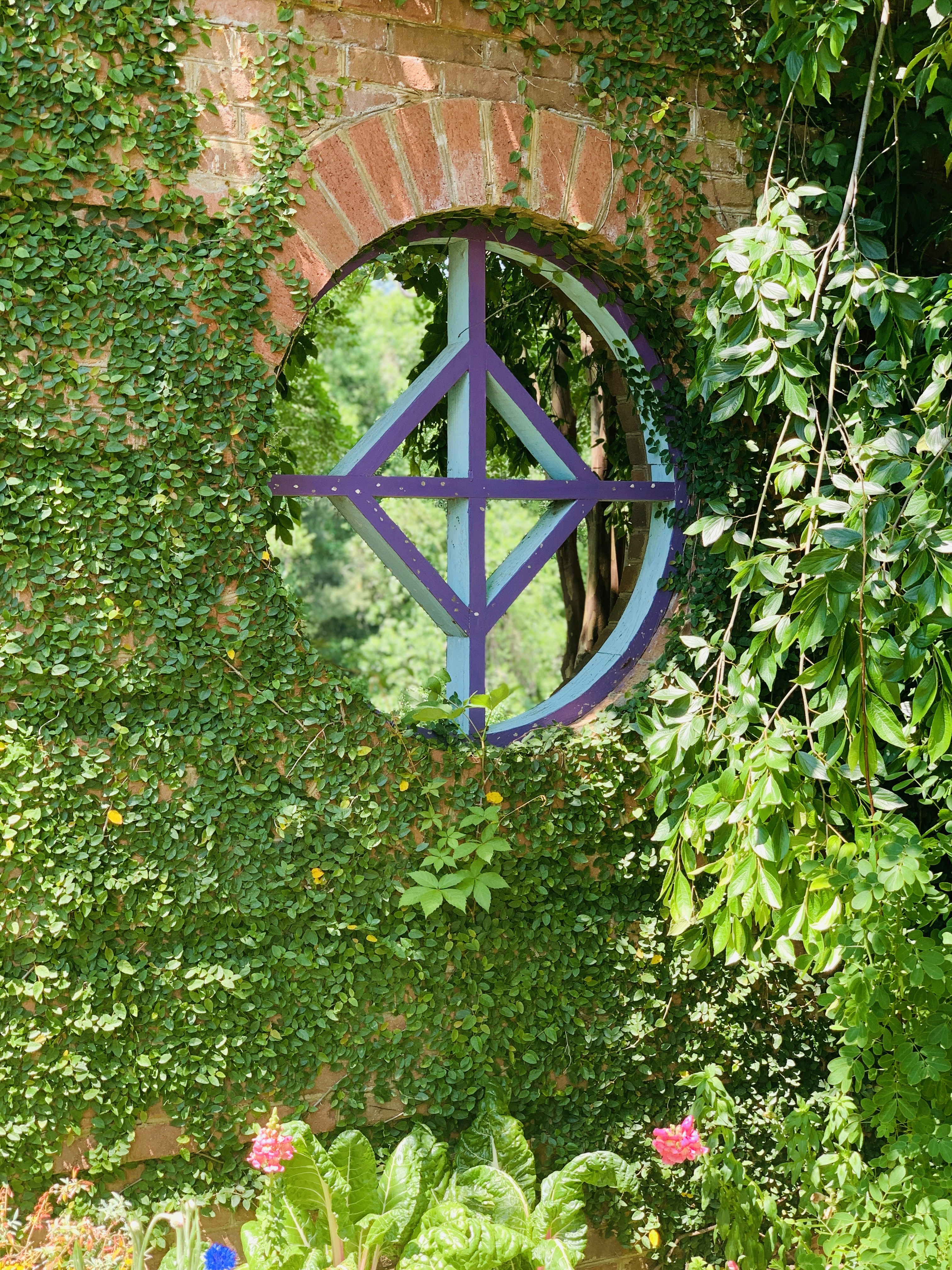 Vibrant purple geometric design framed by lush green ivy on a brick wall, with colorful flowers in the foreground.