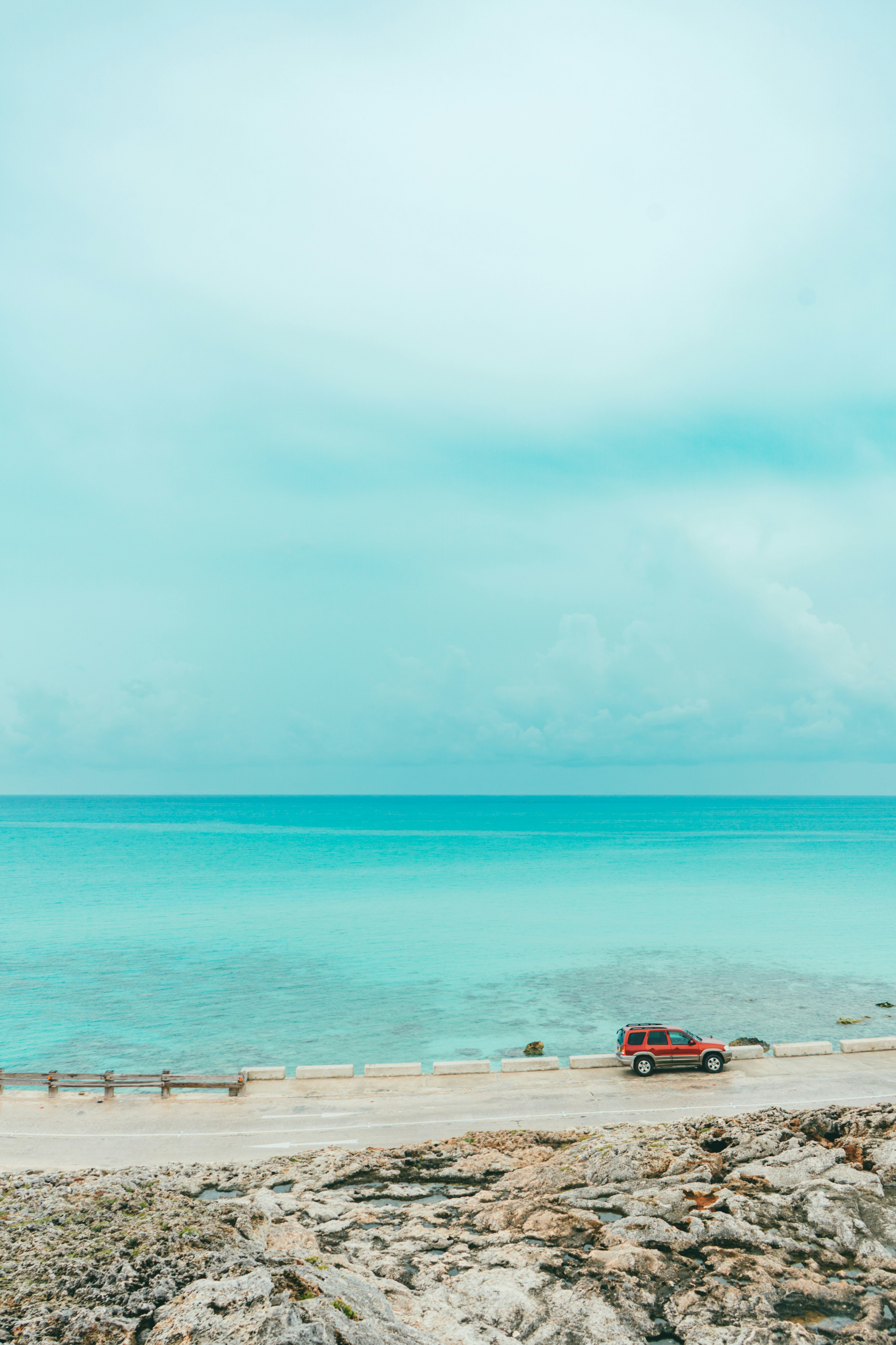 A red vehicle parked along a coastal road, overlooking a tranquil turquoise sea under a cloudy sky.