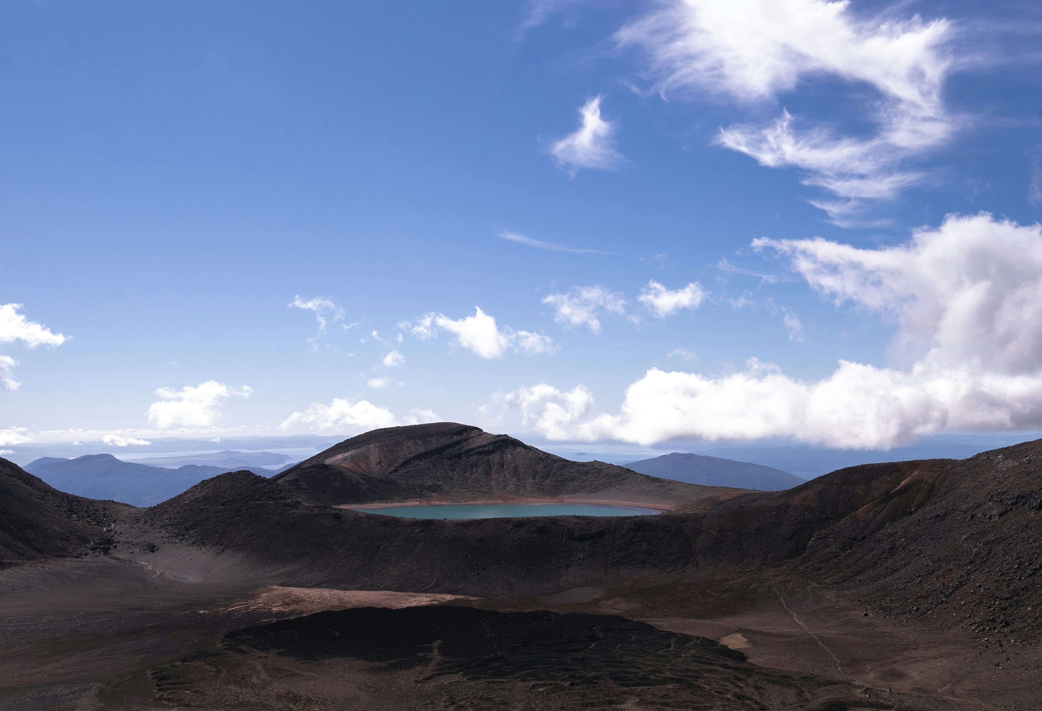 Volcanic landscape with a turquoise crater lake under a vast blue sky with scattered clouds.
