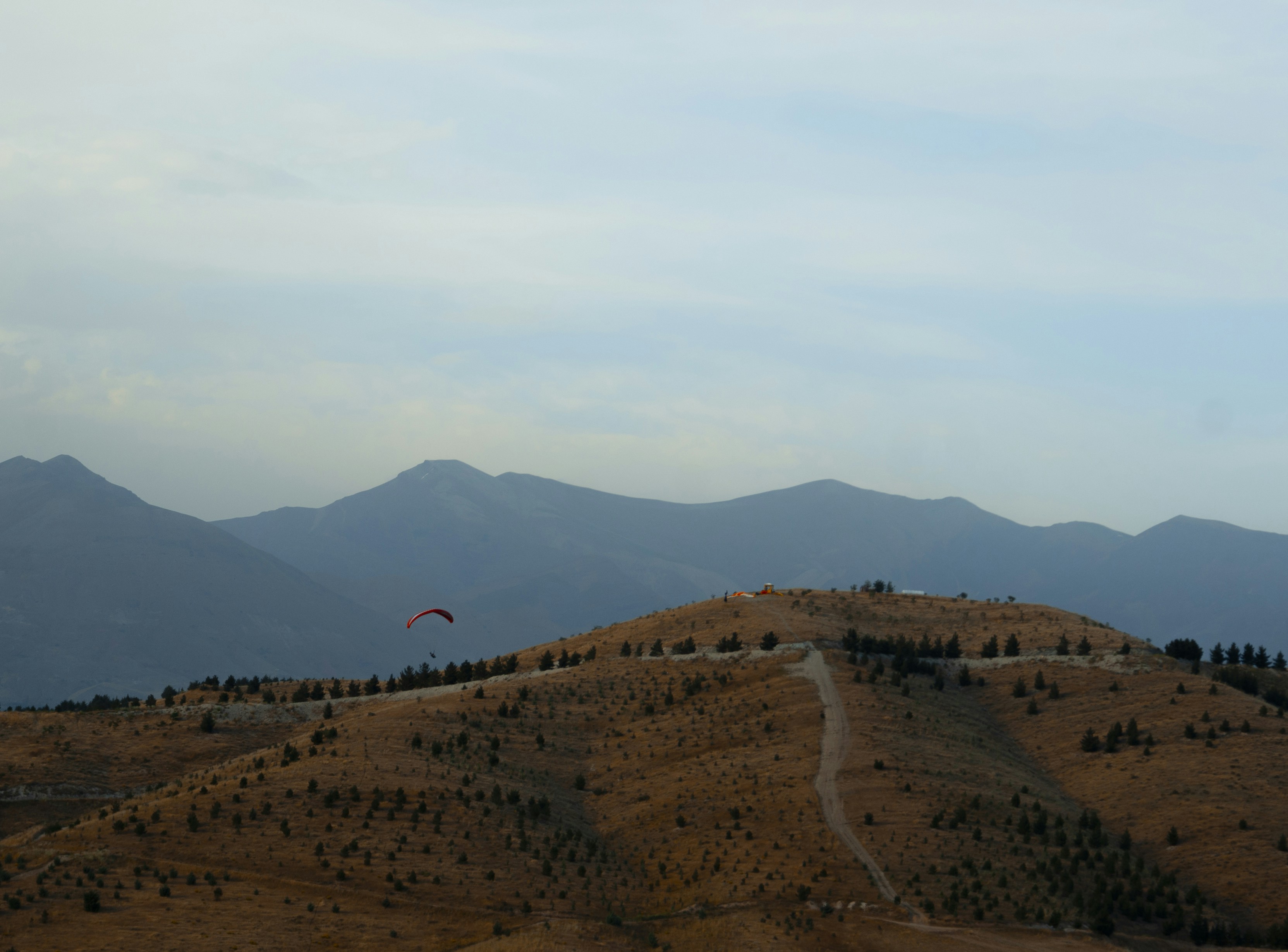 A paraglider gracefully navigates the open skies above rolling golden hills and distant mountain ranges.