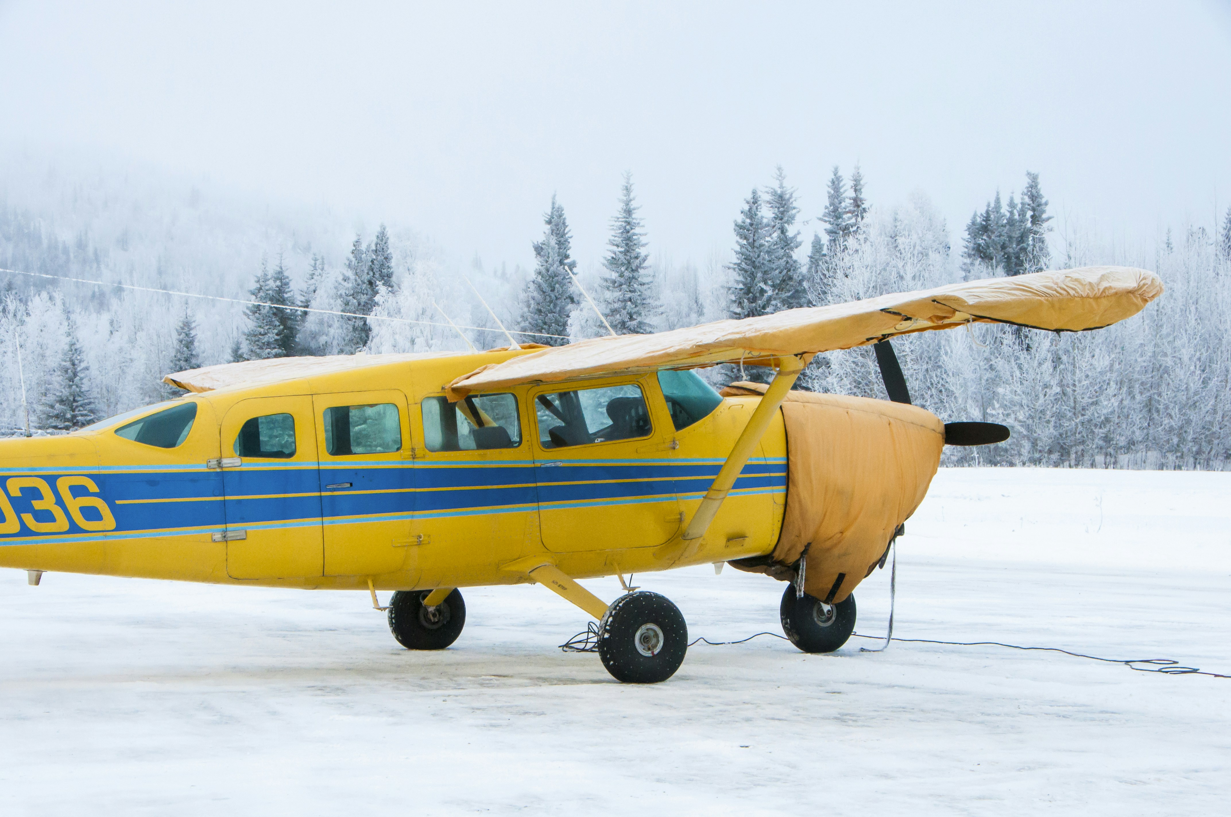 A yellow bush plane parked on a snowy runway, surrounded by frosty trees under a cloudy sky.