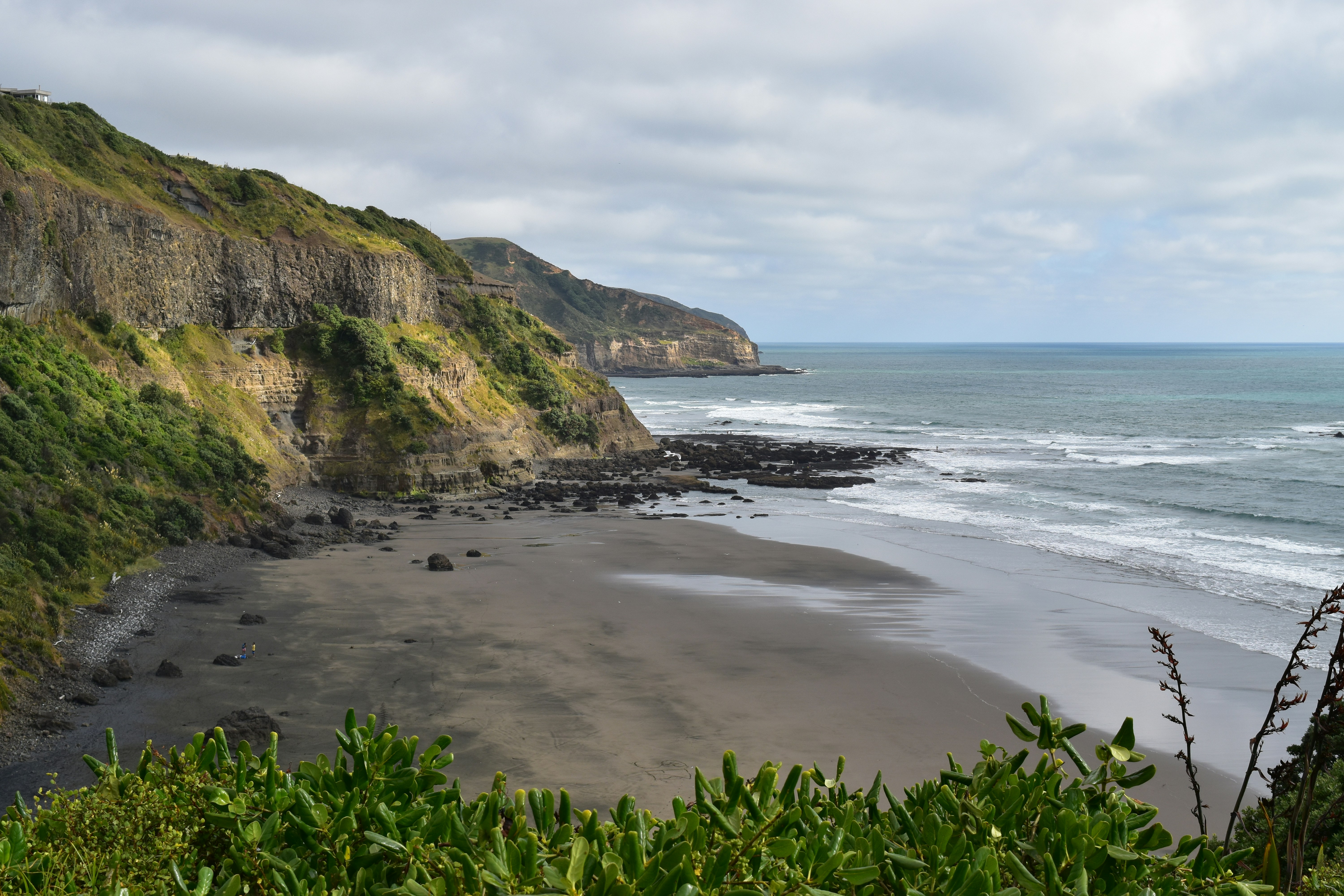Rocky cliffs meet the ocean, with waves gently lapping at a secluded sandy beach under a cloudy sky.