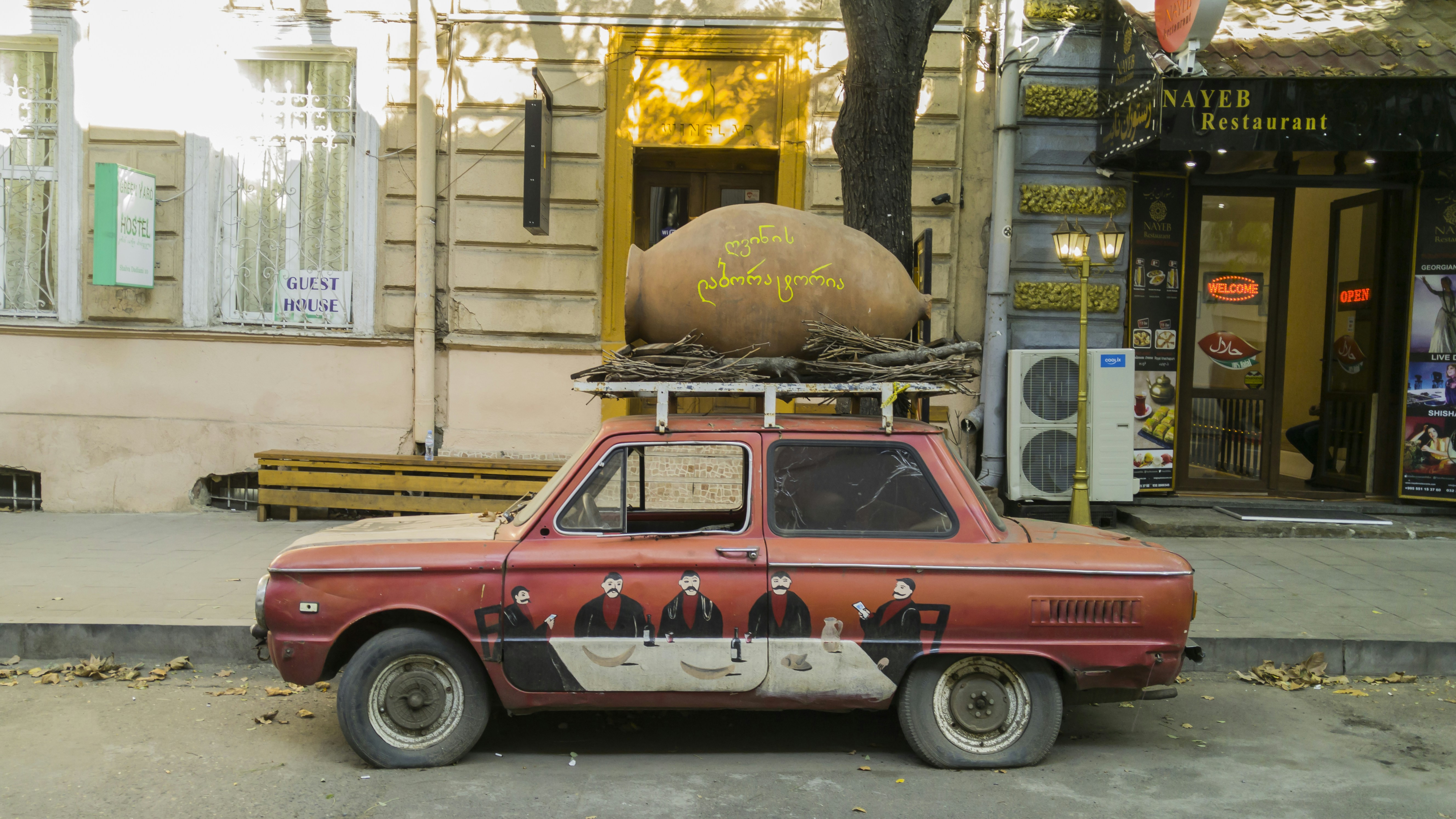 A vintage car adorned with a mural of local figures, topped with a large clay pot, parked outside a restaurant in a charming urban setting.