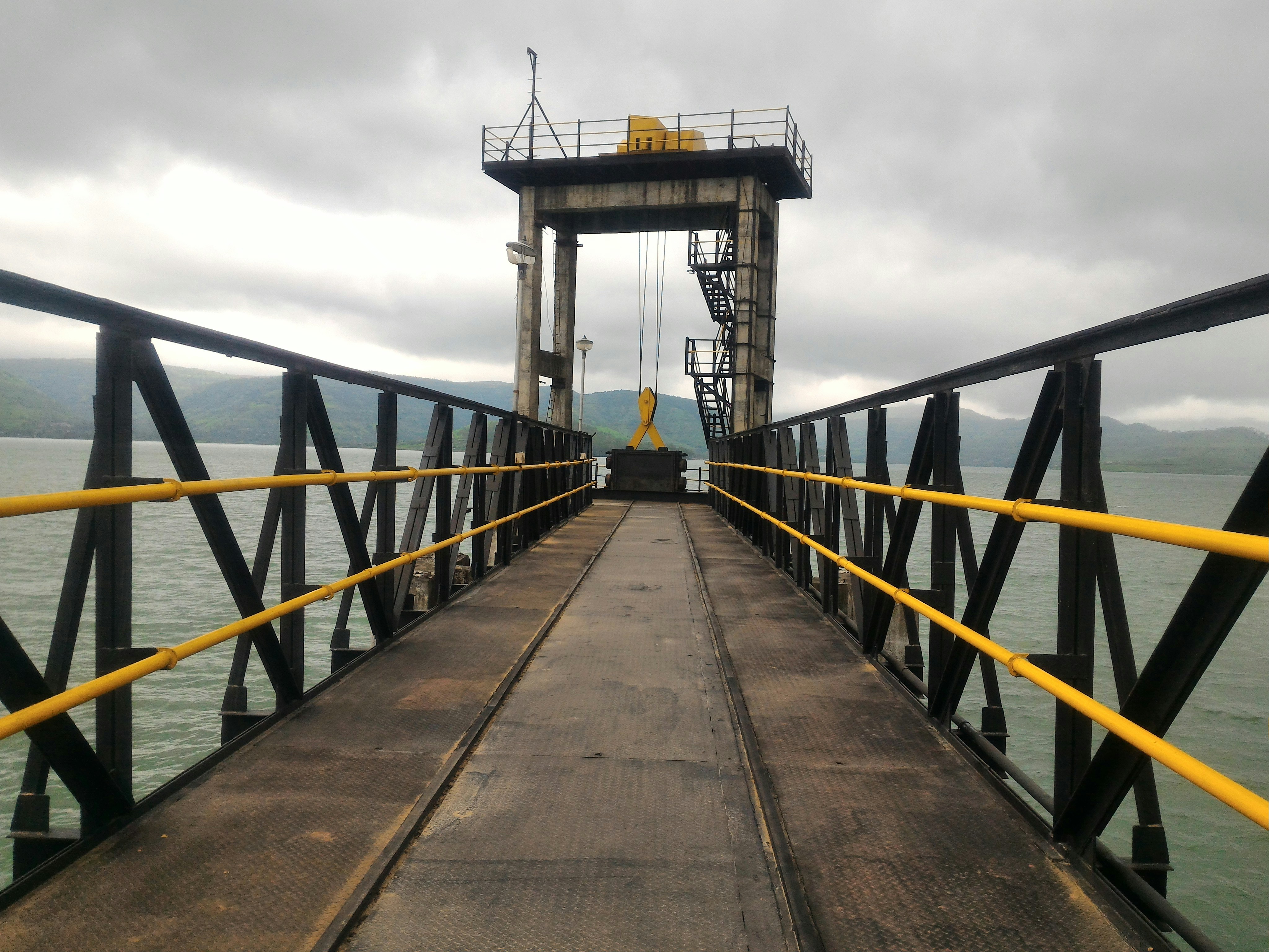 Rusted steel pier walkway with yellow handrails extends toward a distant control tower over gray water. Overcast sky adds a moody atmosphere.