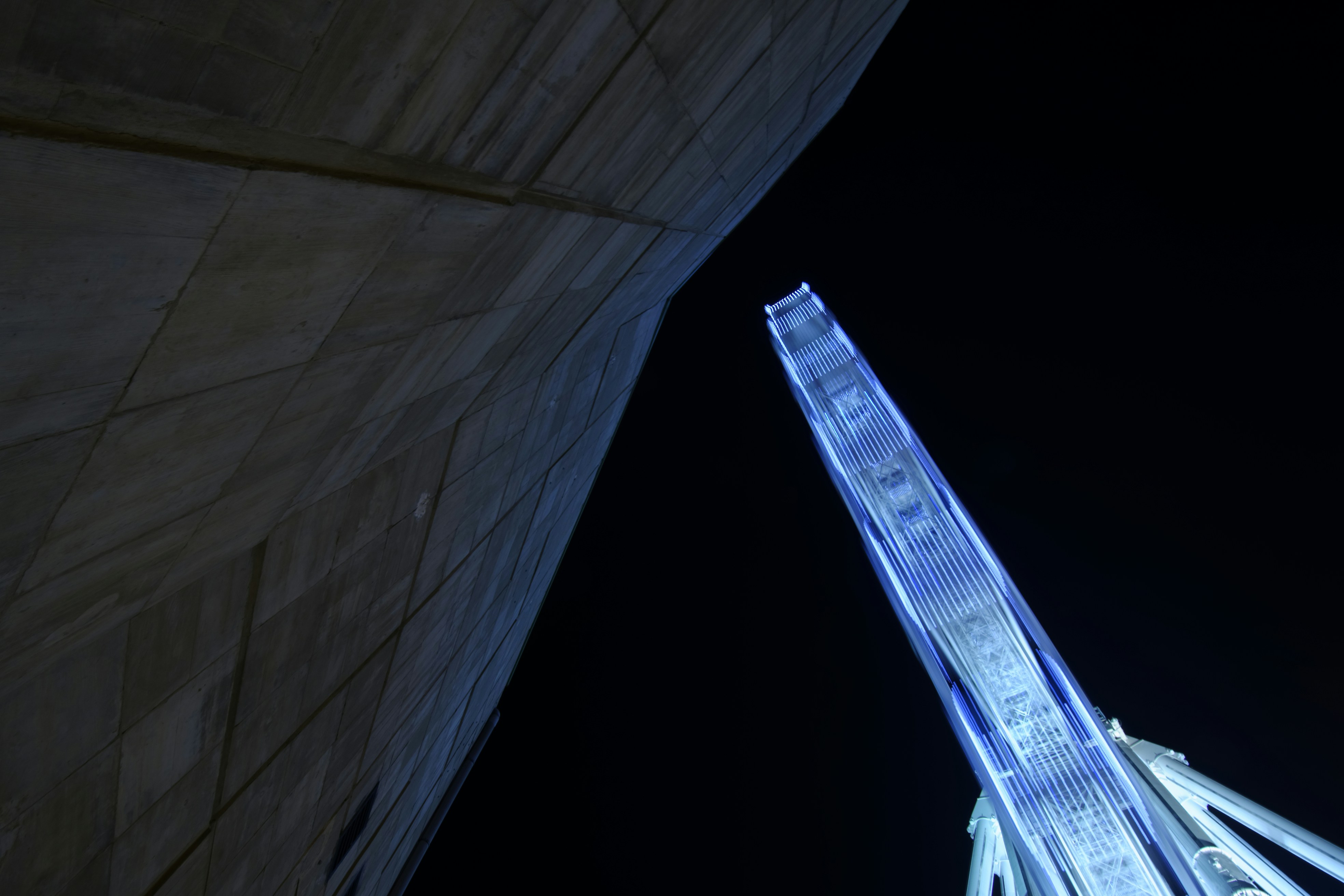 A striking tower illuminated in blue reaches skyward, juxtaposed against a textured concrete wall at night.