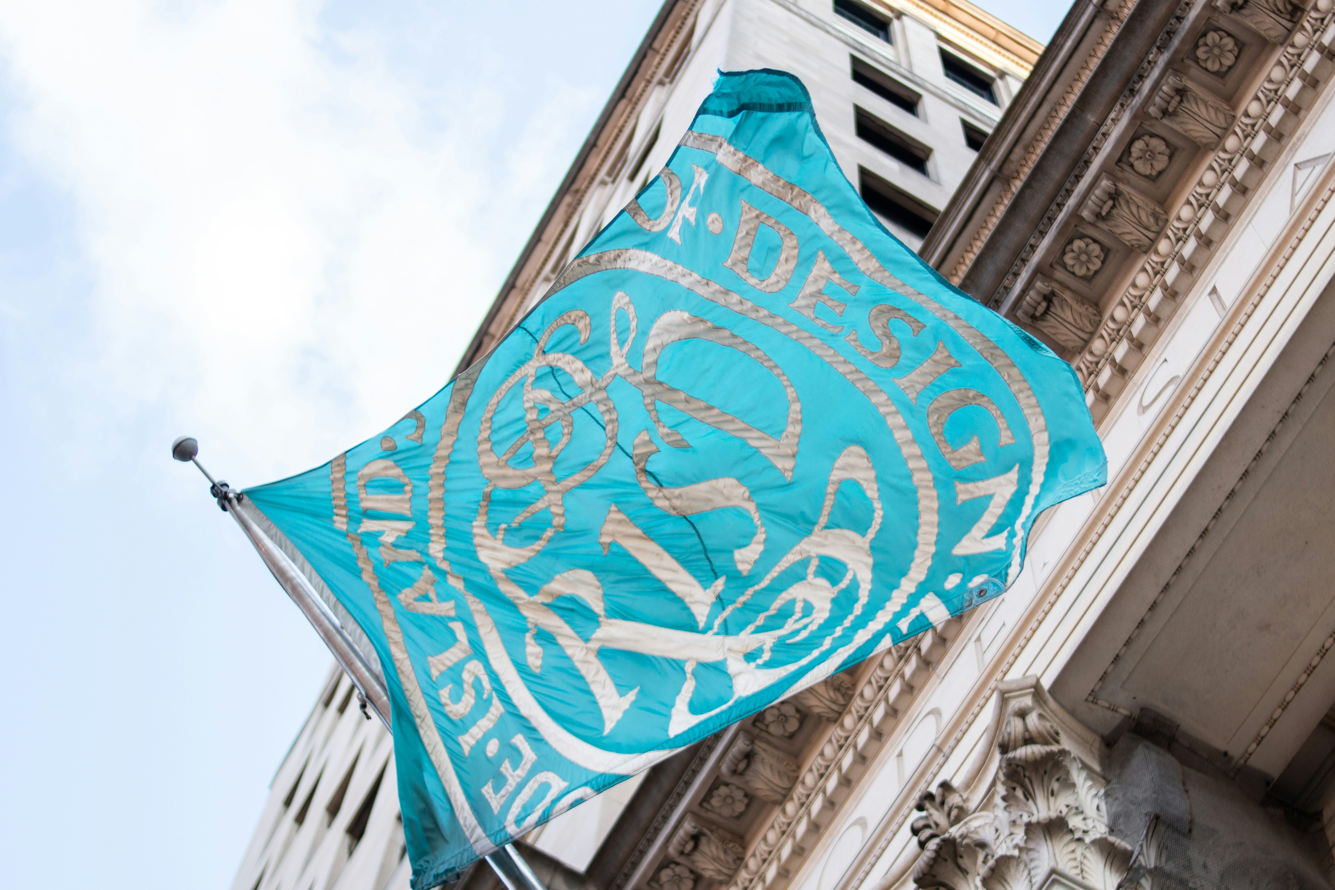 Ornate teal flag with emblem, flying against a backdrop of a historic building facade and cloudy sky.